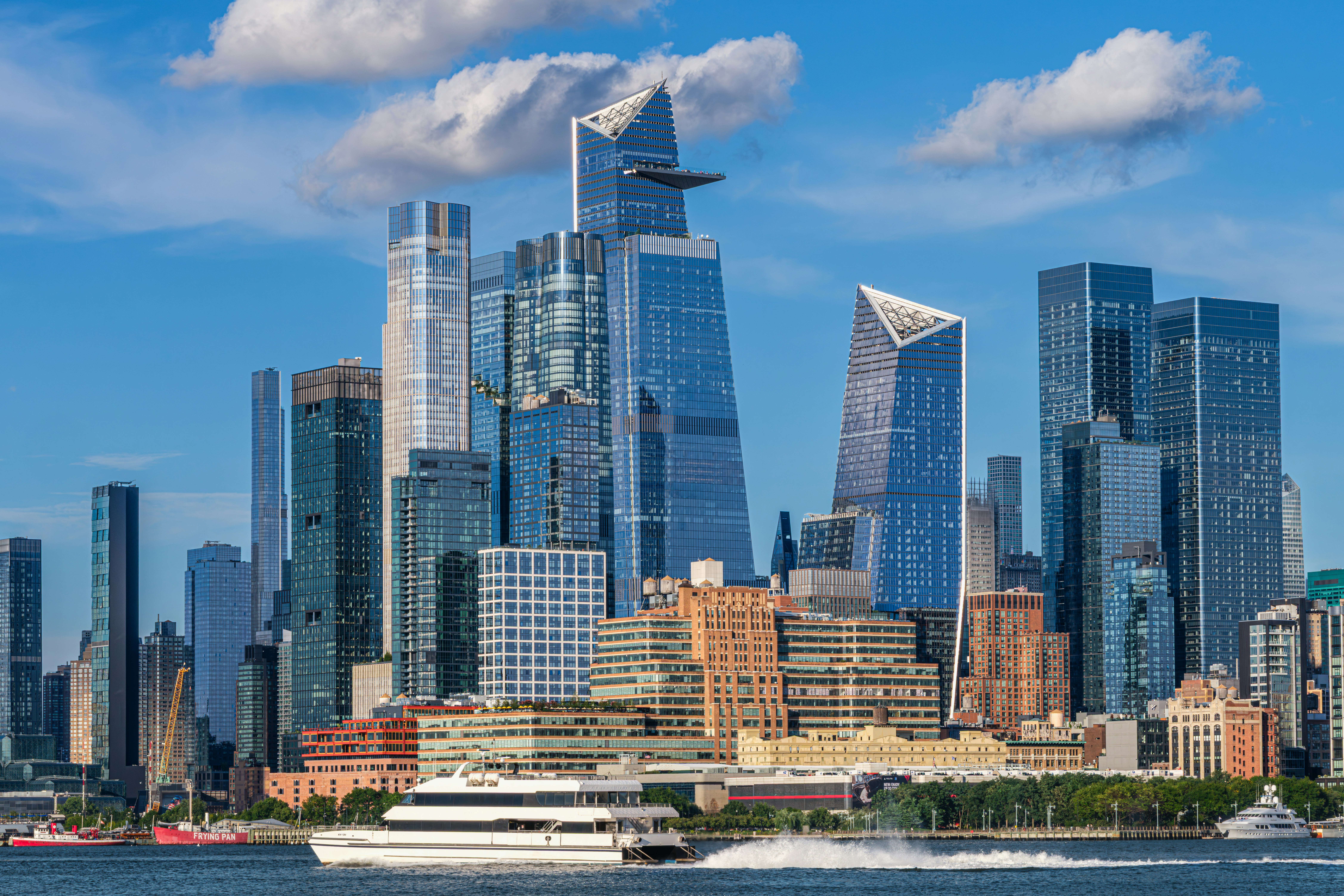 A modern city skyline rises along a calm harbor with a white passenger boat passing in the foreground; glass towers reflect the blue sky, displaying a mix of tall skyscrapers and mid-rise structures.