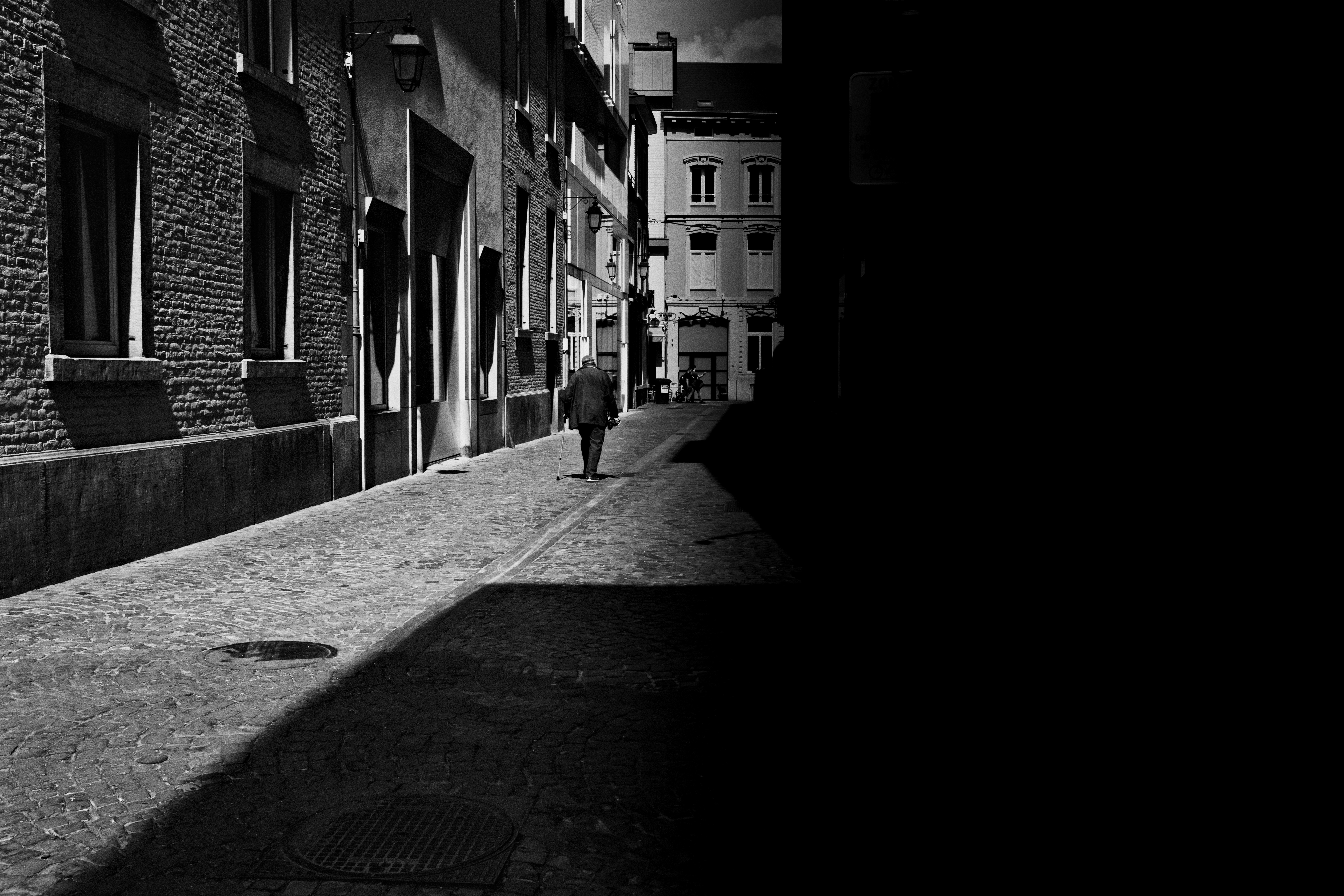 A person walking with a cane down a narrow cobblestone alley in high contrast light