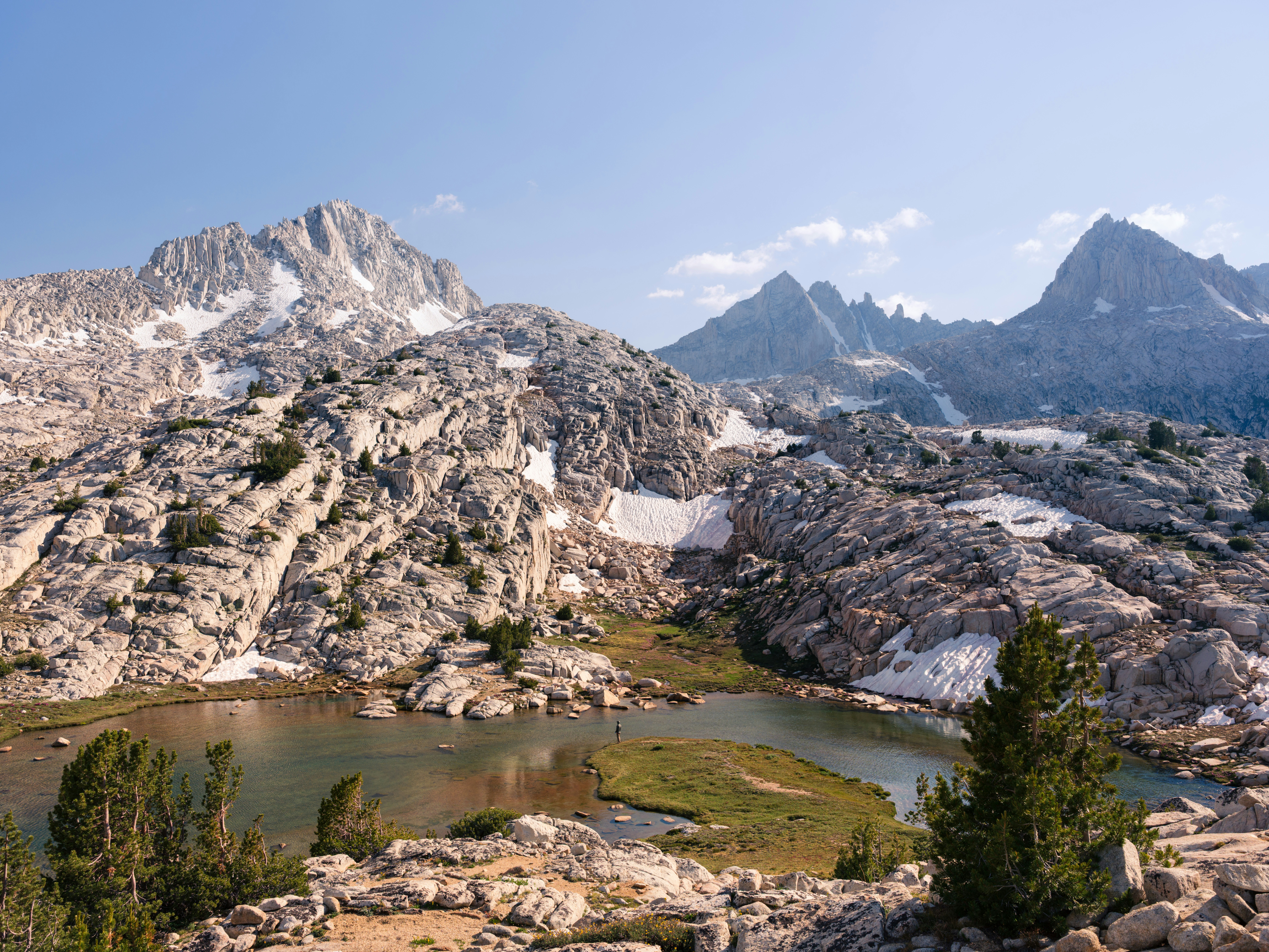 Snow-dusted peaks rise over a rocky basin with a small pond and scattered evergreens, under a clear blue sky.
