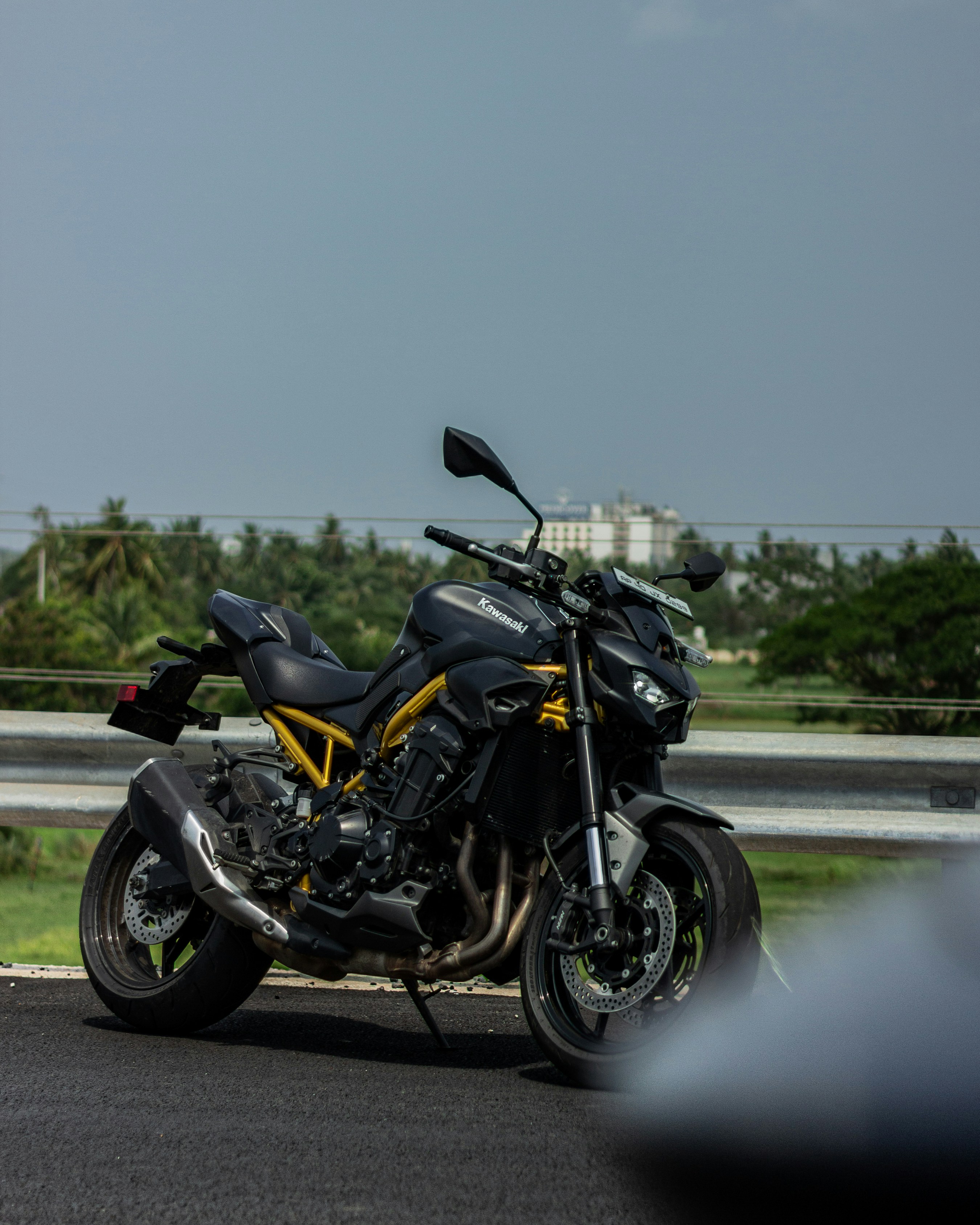 Dark Kawasaki motorcycle parked on a road with guardrail and distant buildings, under soft daylight; the bike dominates the frame with focus on its angular frame and exhaust. This is a photographic capture of a street machine scene.