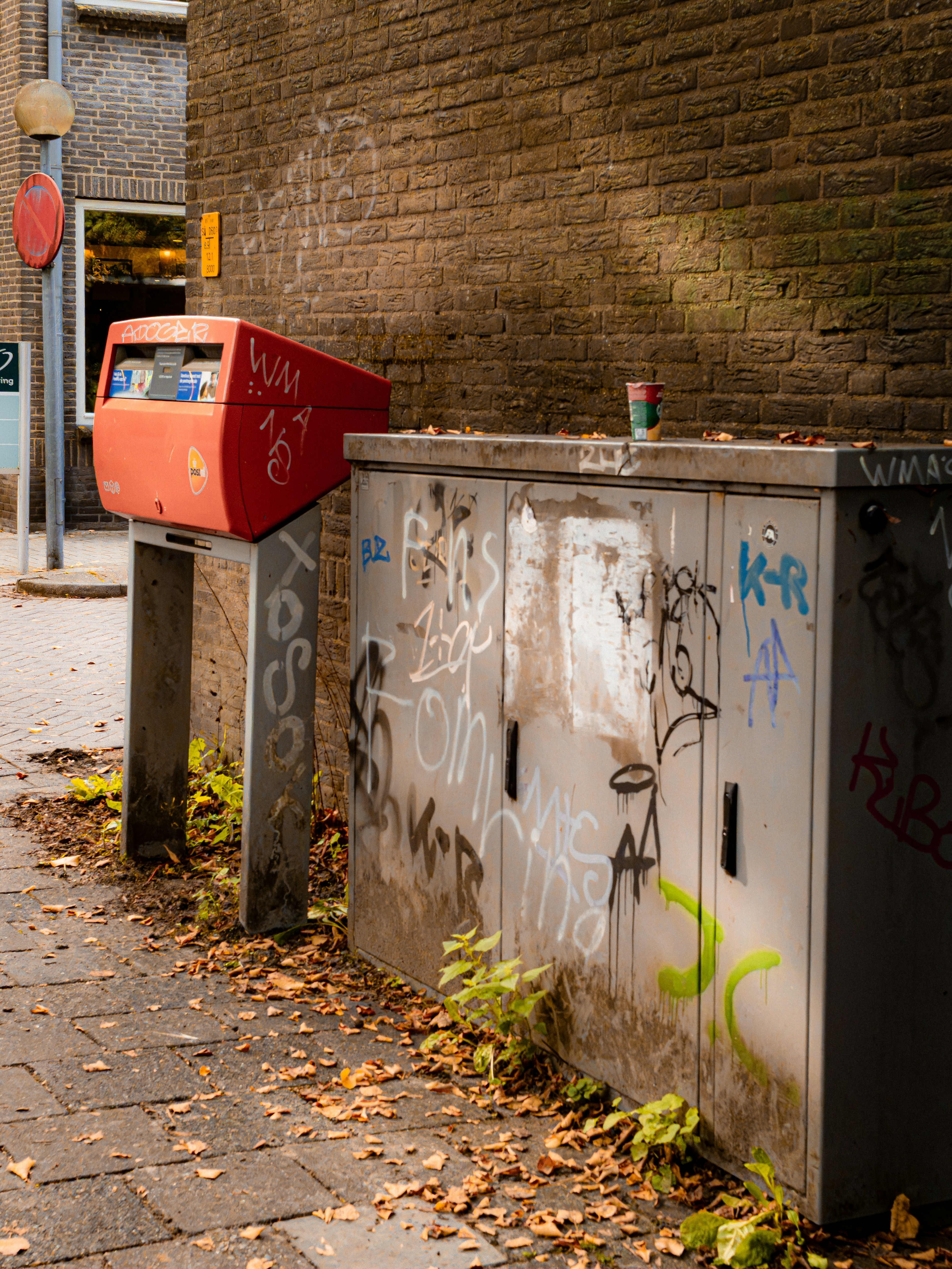Red mailbox perched beside a graffiti-streaked utility cabinet along a city sidewalk; autumn leaves scatter the ground, a yellow curb sign peeks from behind, and a brick wall provides a warm backdrop.