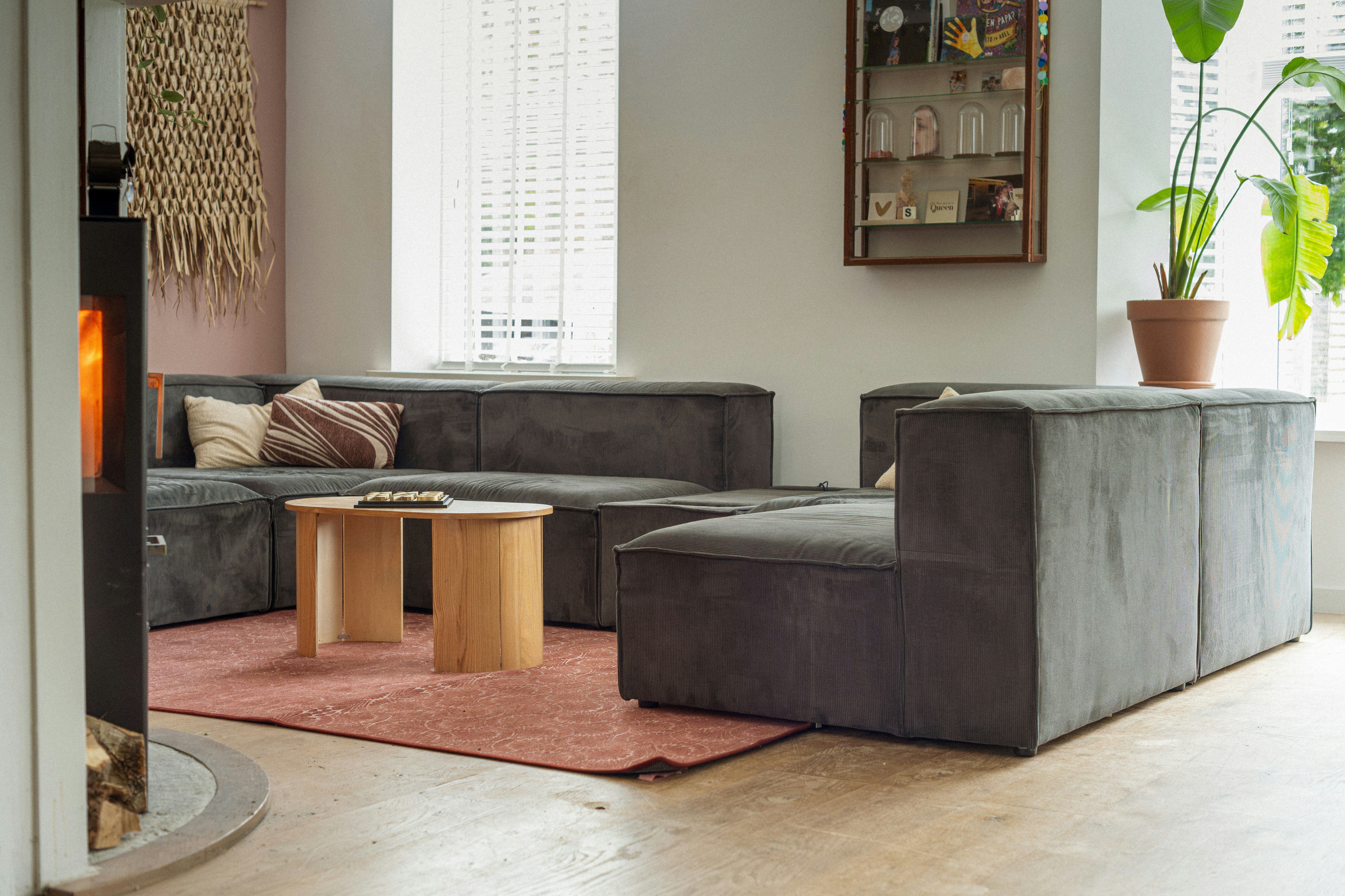 A sunlit living room featuring a dark sectional, a round wooden coffee table, and a woven rug, with a fireplace to the left and houseplants adding warmth.