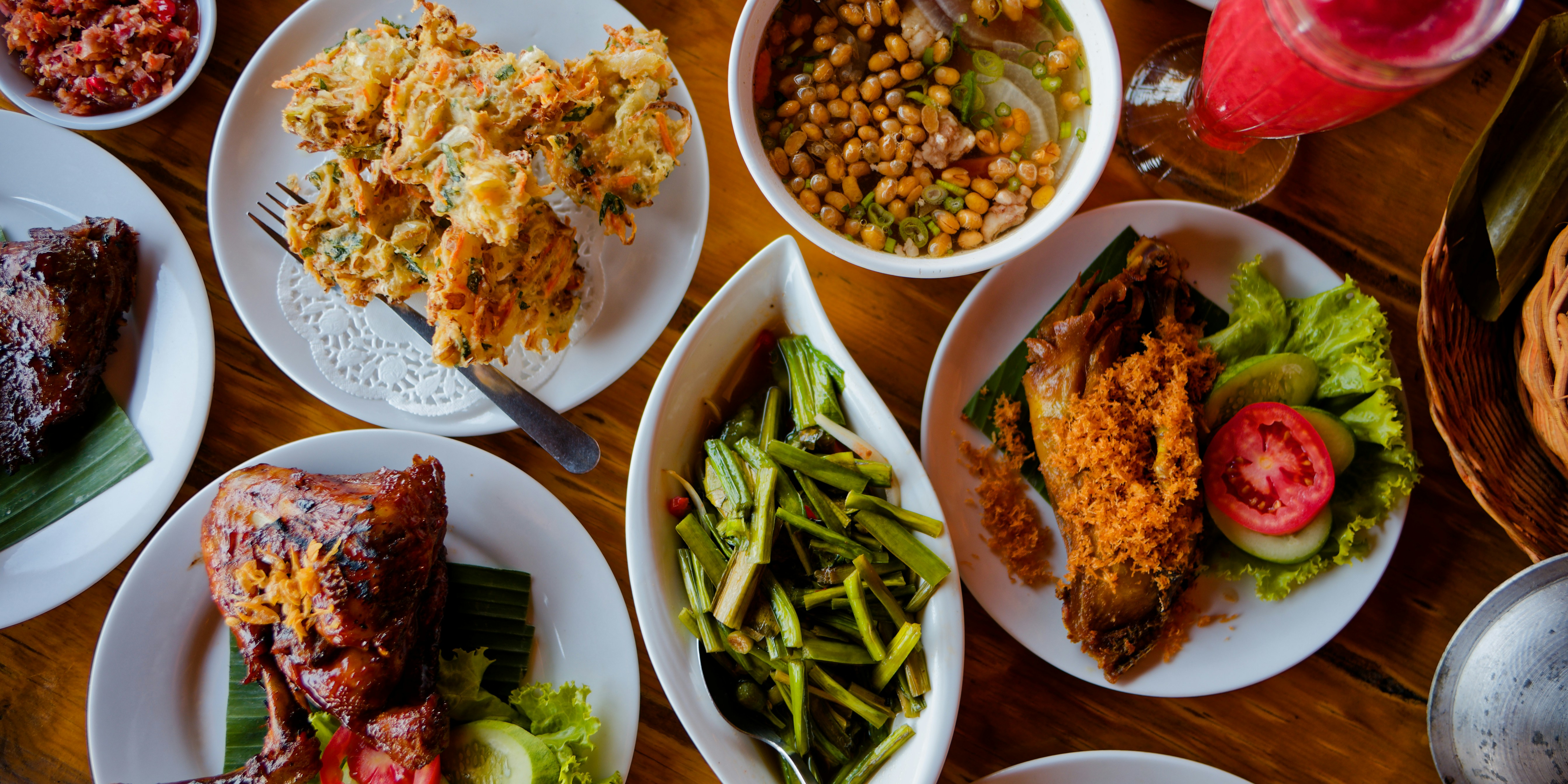 Colorful array of plated dishes on a wooden table, featuring fried items, greens, legumes, and a pink beverage; a communal meal setting highlighting varied textures and flavors.