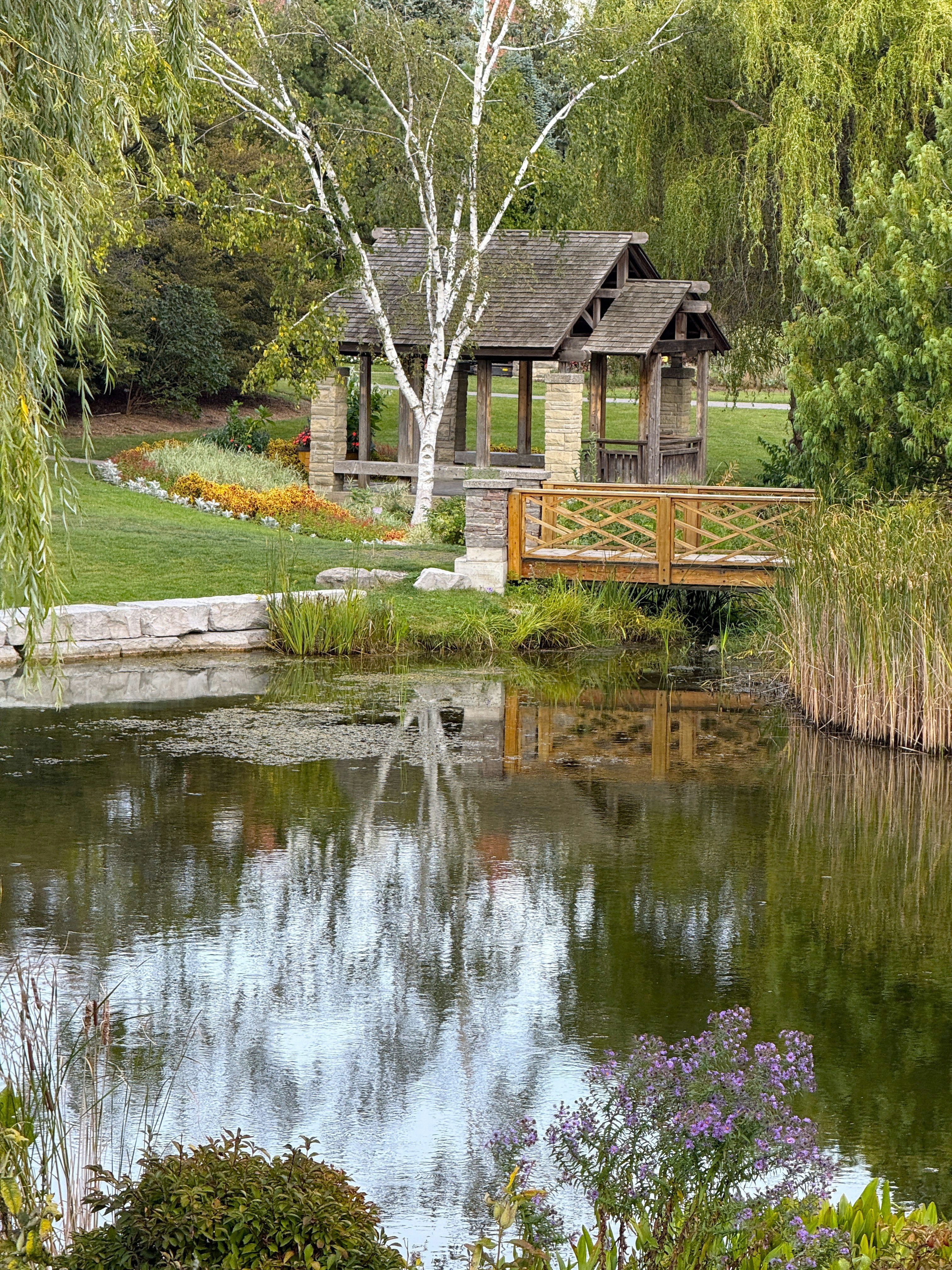 Quiet garden scene with a wooden gazebo over a small bridge spanning a reflective pond, surrounded by greenery and flowering borders.
