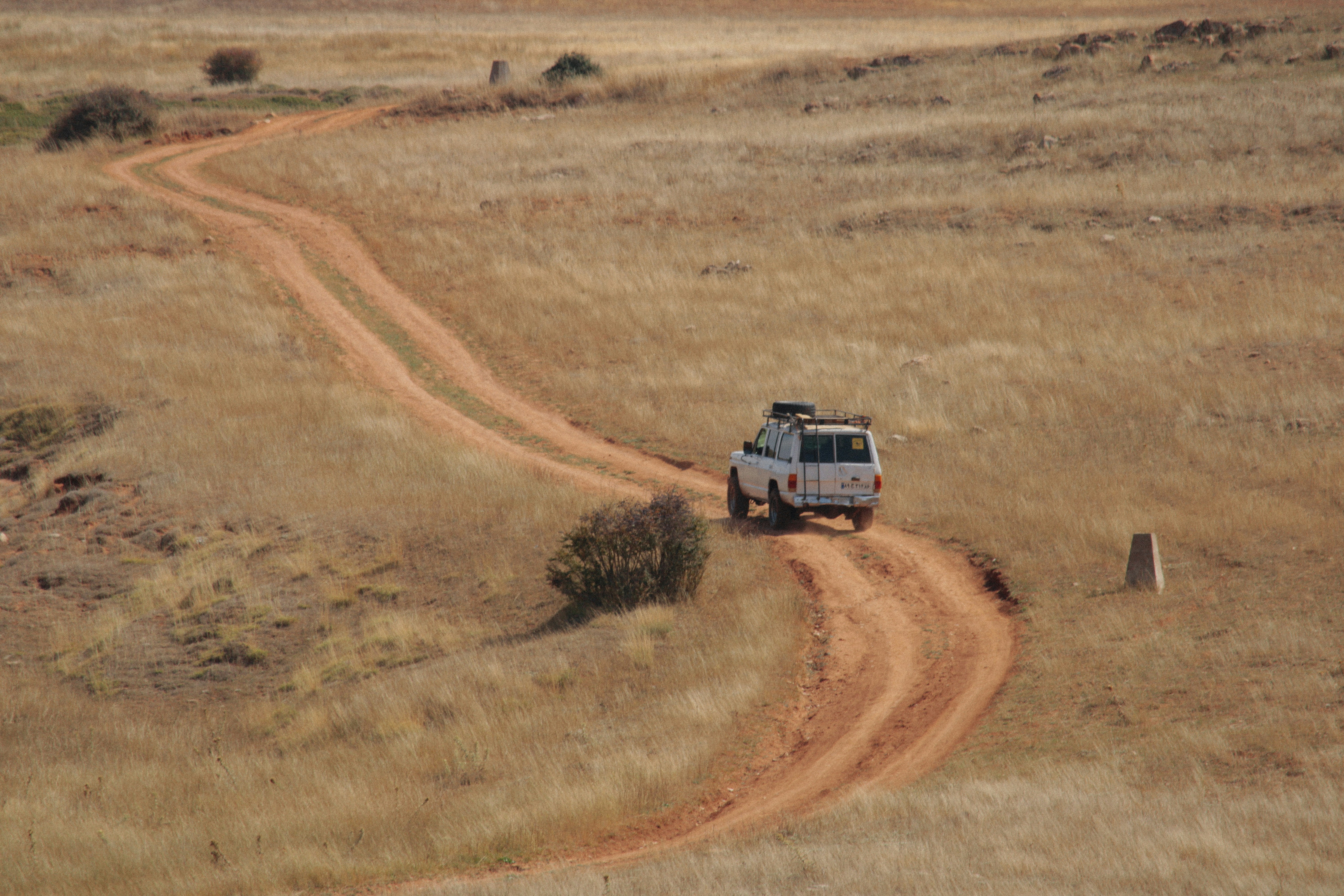 Dusty white SUV on a winding dirt track through arid grassland under muted light.