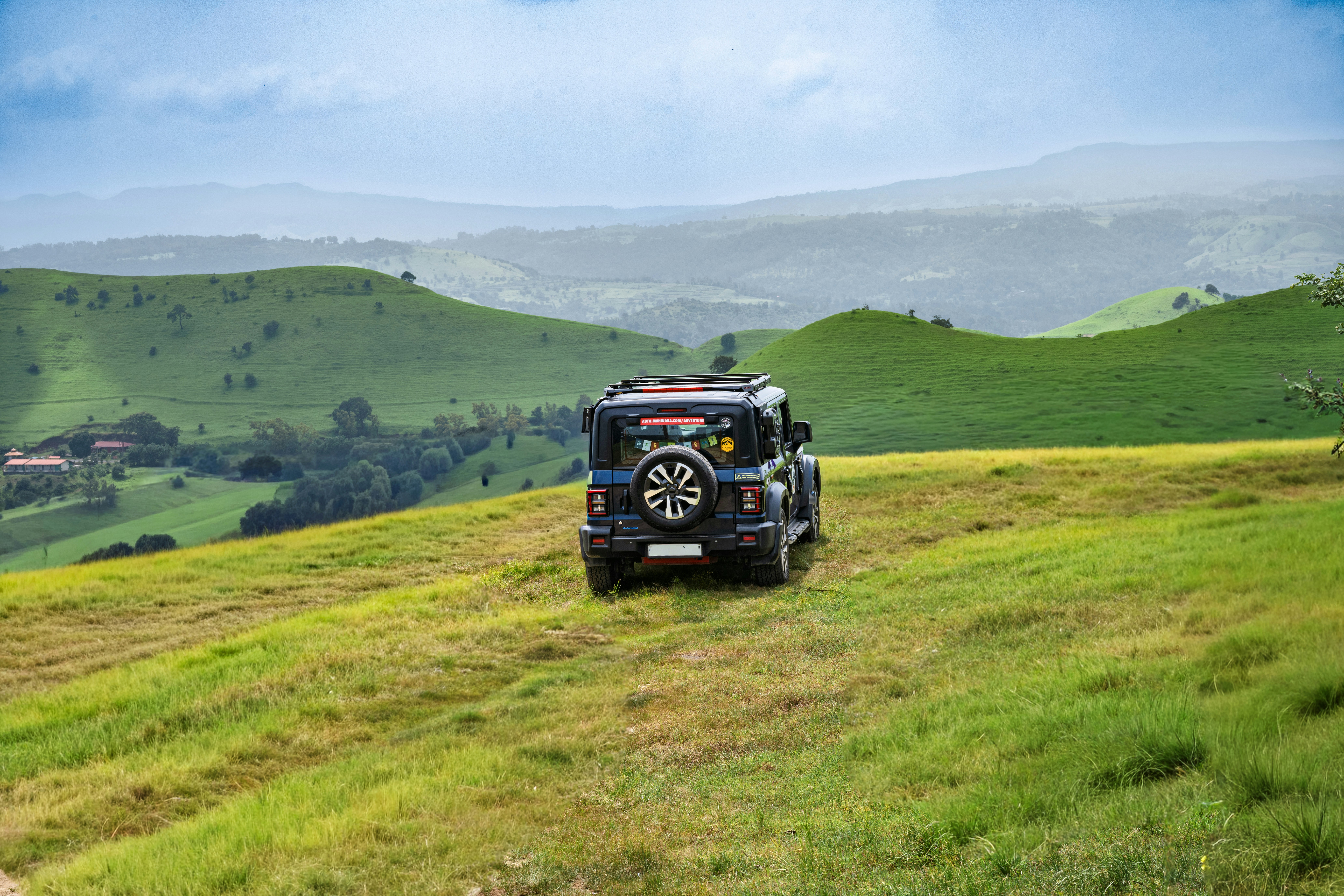 Off-road jeep on a grassy hillside with rolling green valleys and distant mountains under a blue sky. A landscape shot highlighting the vehicle as the focal point in a wide, open setting.