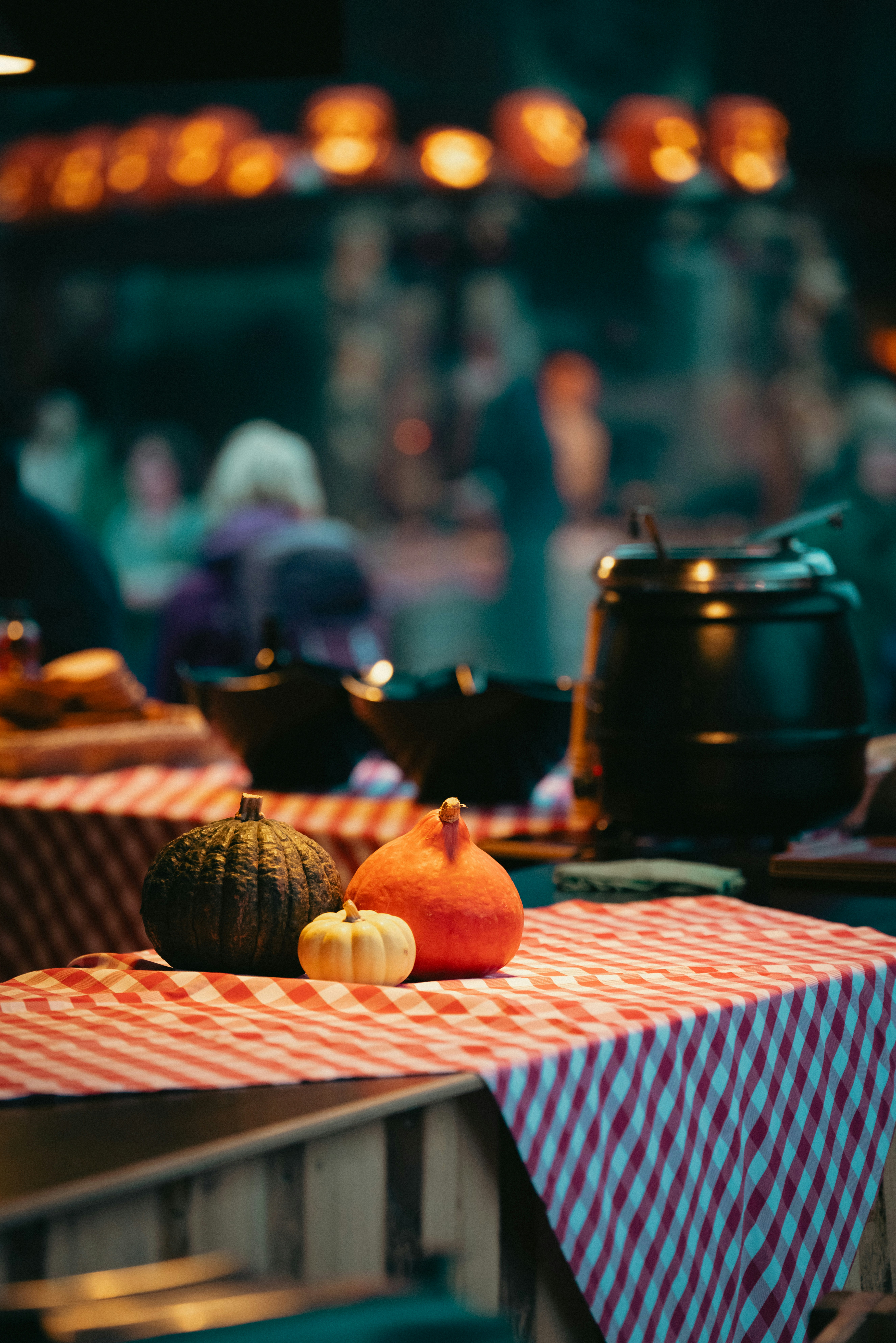 Small pumpkins rest on a checkered tablecloth at a warm, blurred market scene with a slow-cooked pot in the background.