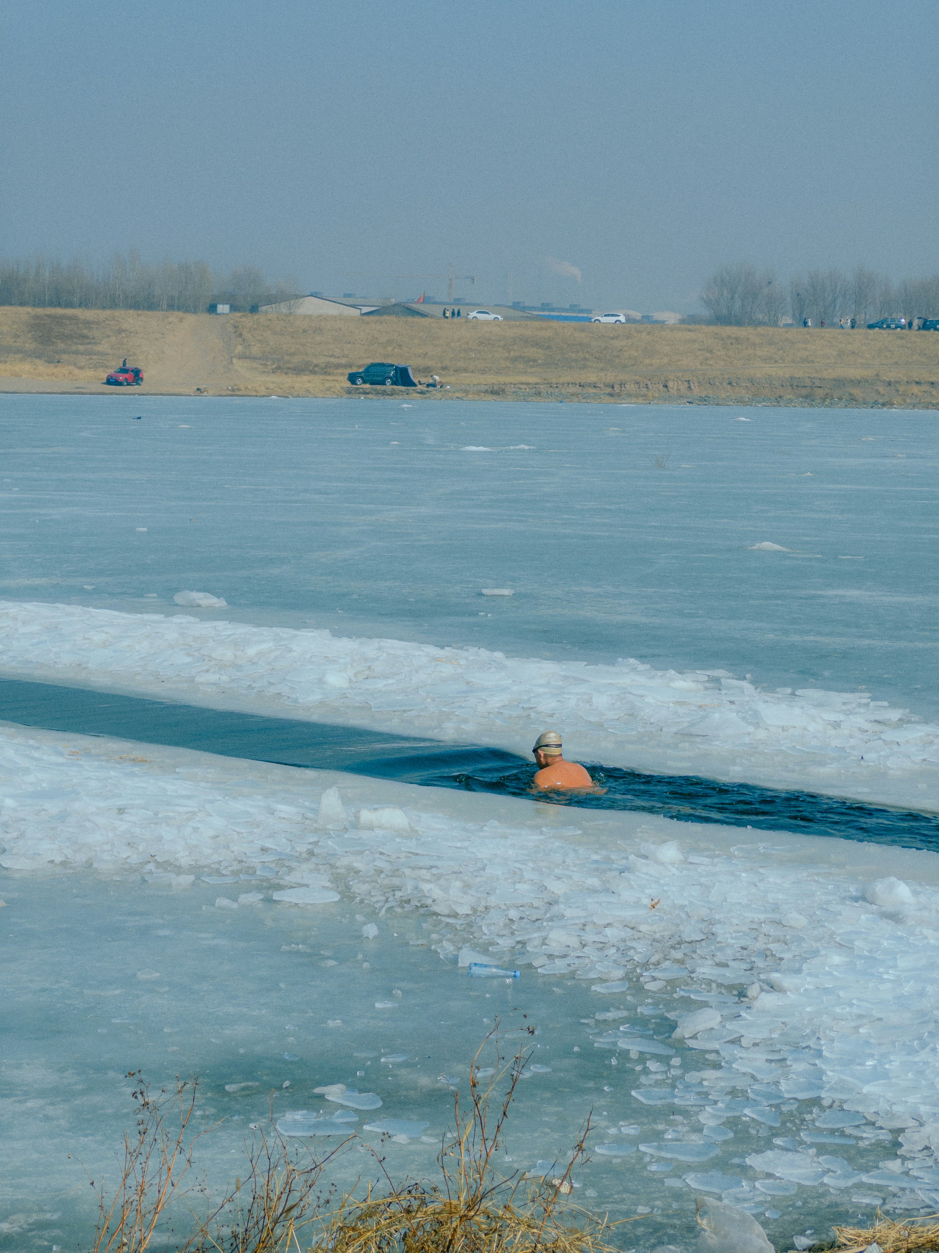 A lone swimmer in an orange suit glides through a fragmented, frozen lake with a distant shoreline and cars in the background.