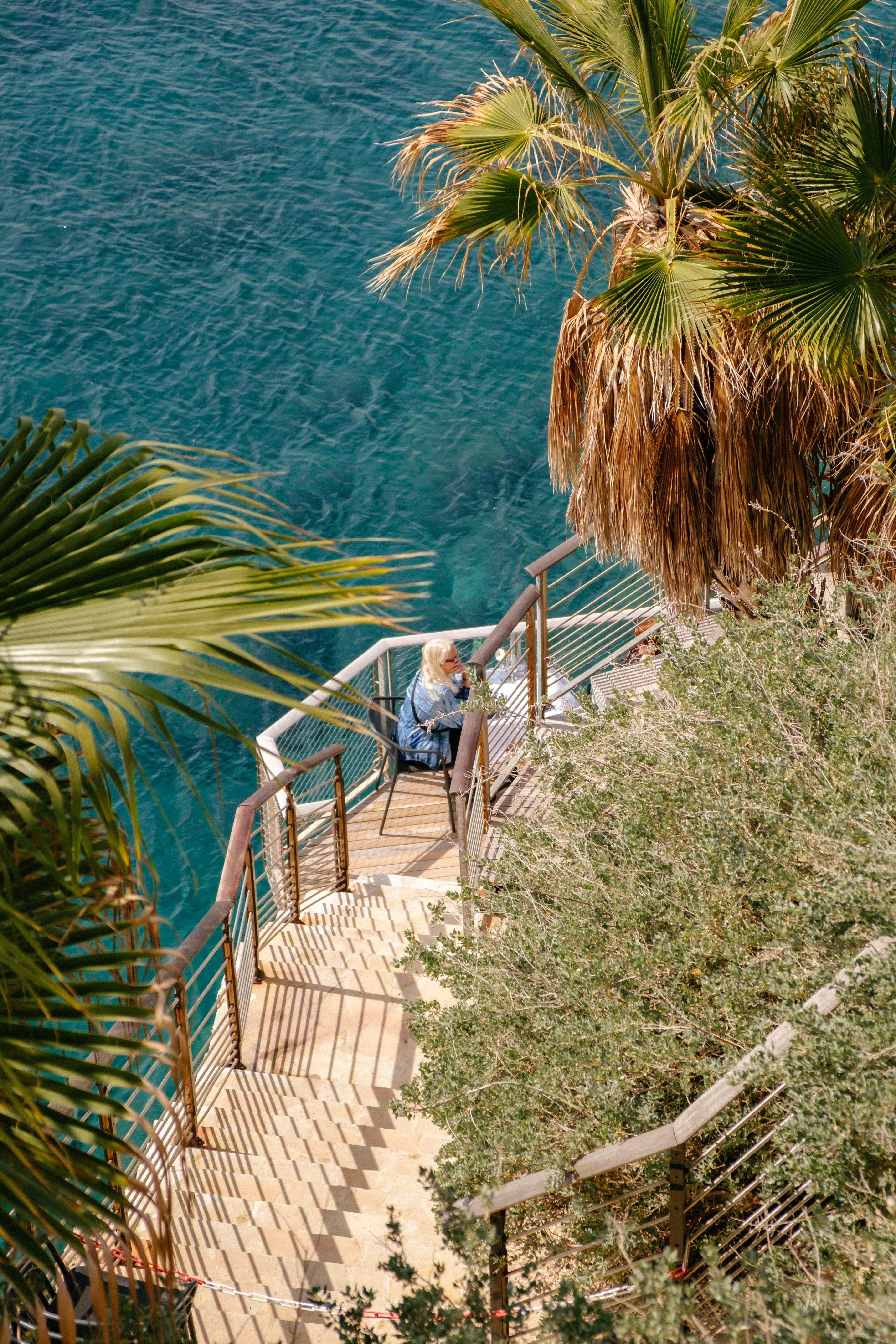 Stairway to Aegean Quiet: A Couple Amid Palm Shadows Over Turquoise Waters