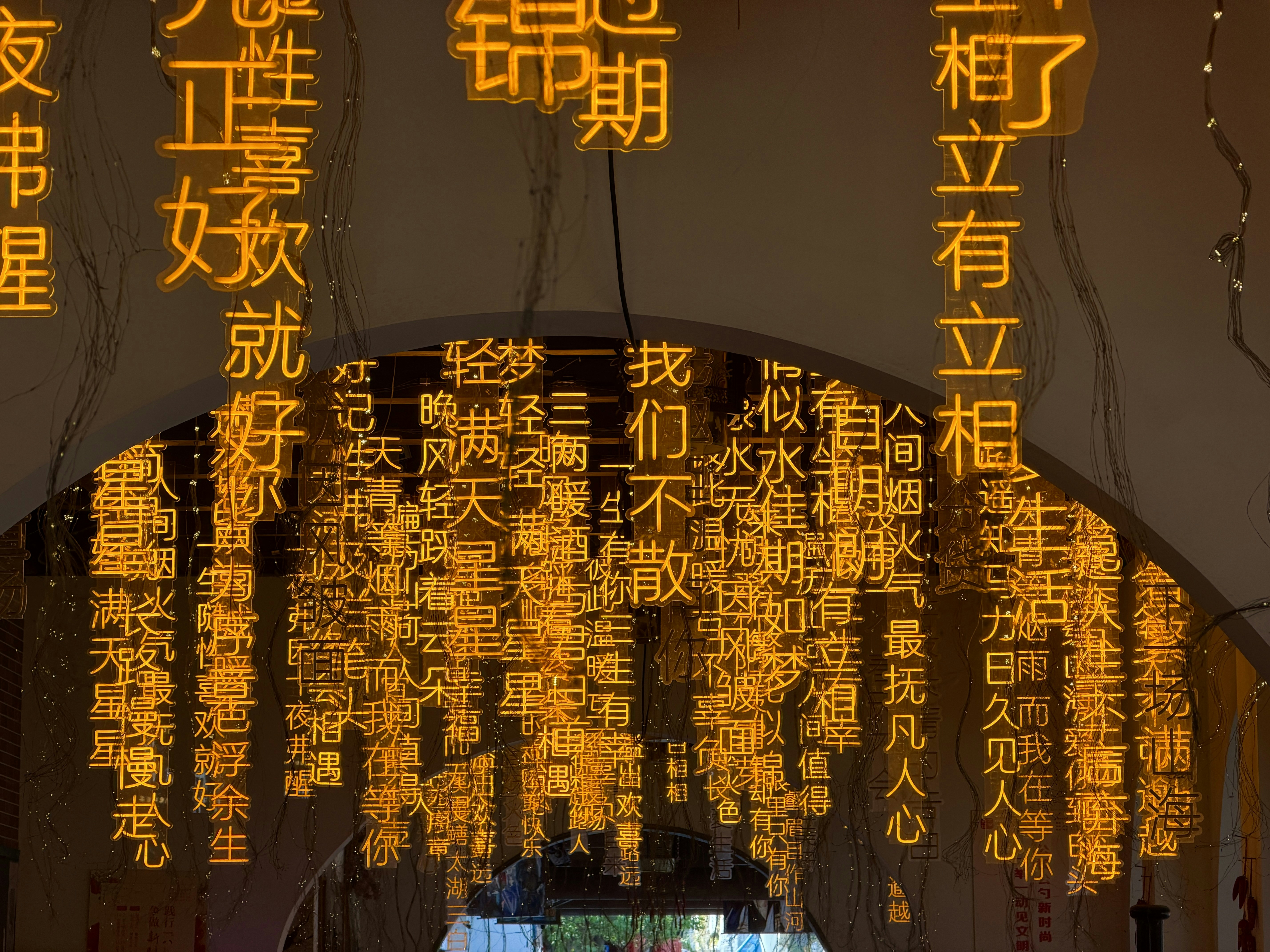 Neon orange script bends into a circular lantern-like display, filling a dark space with warm glow and geometric repetition. The scene emphasizes illuminated characters and their reflections on curved surfaces.