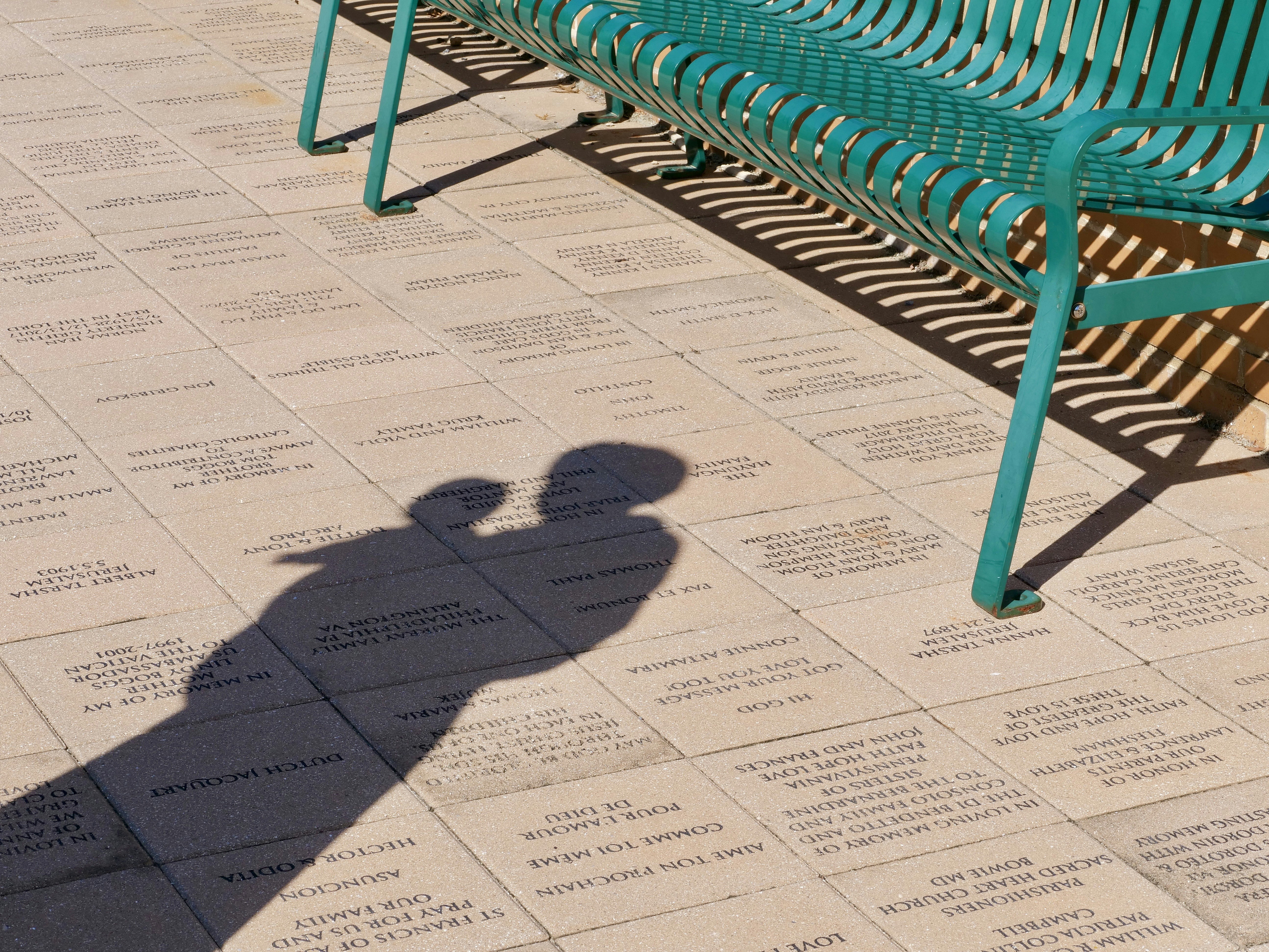 Shadows of two people at a turquoise bench cast over engraved stone tiles, suggesting a quiet moment in an outdoor plaza.