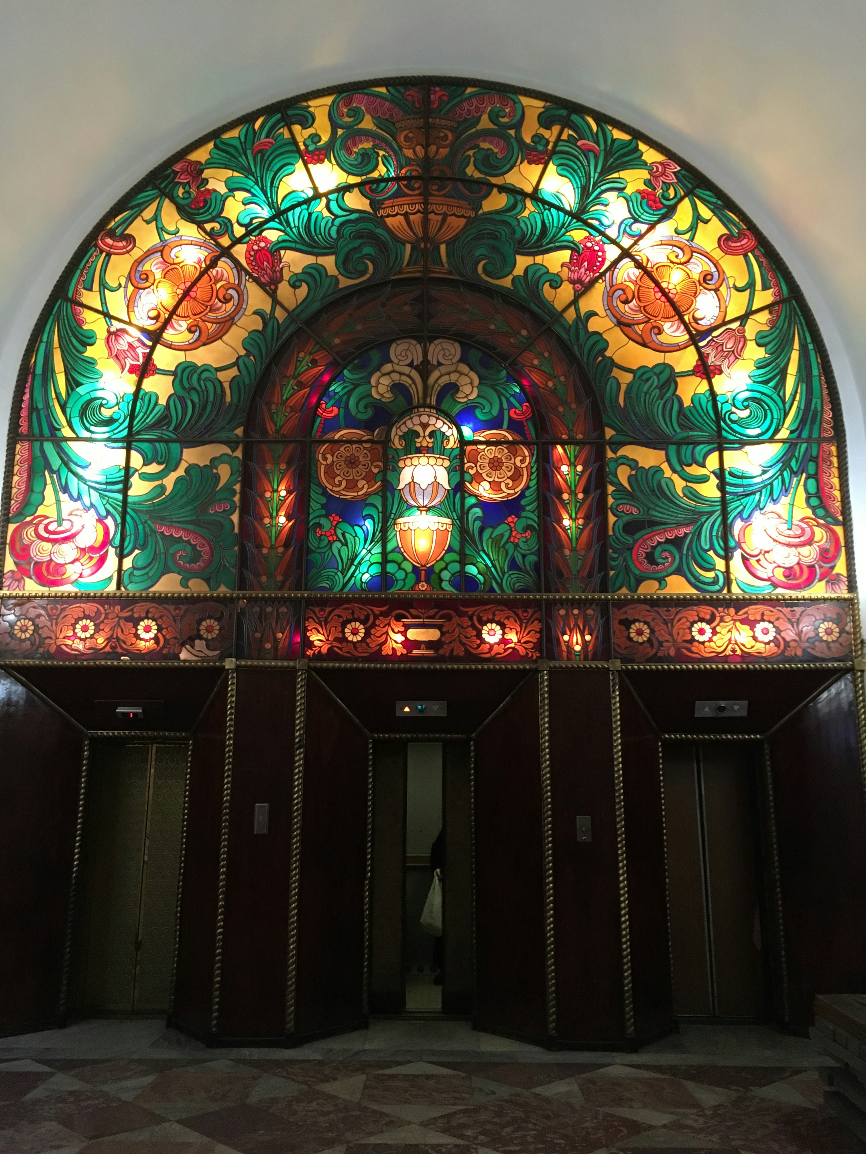 Colorful stained-glass arch over illuminated elevator bays in a dim lobby; ornate panels and mosaic floor anchor a ceremonial, decorative setting.