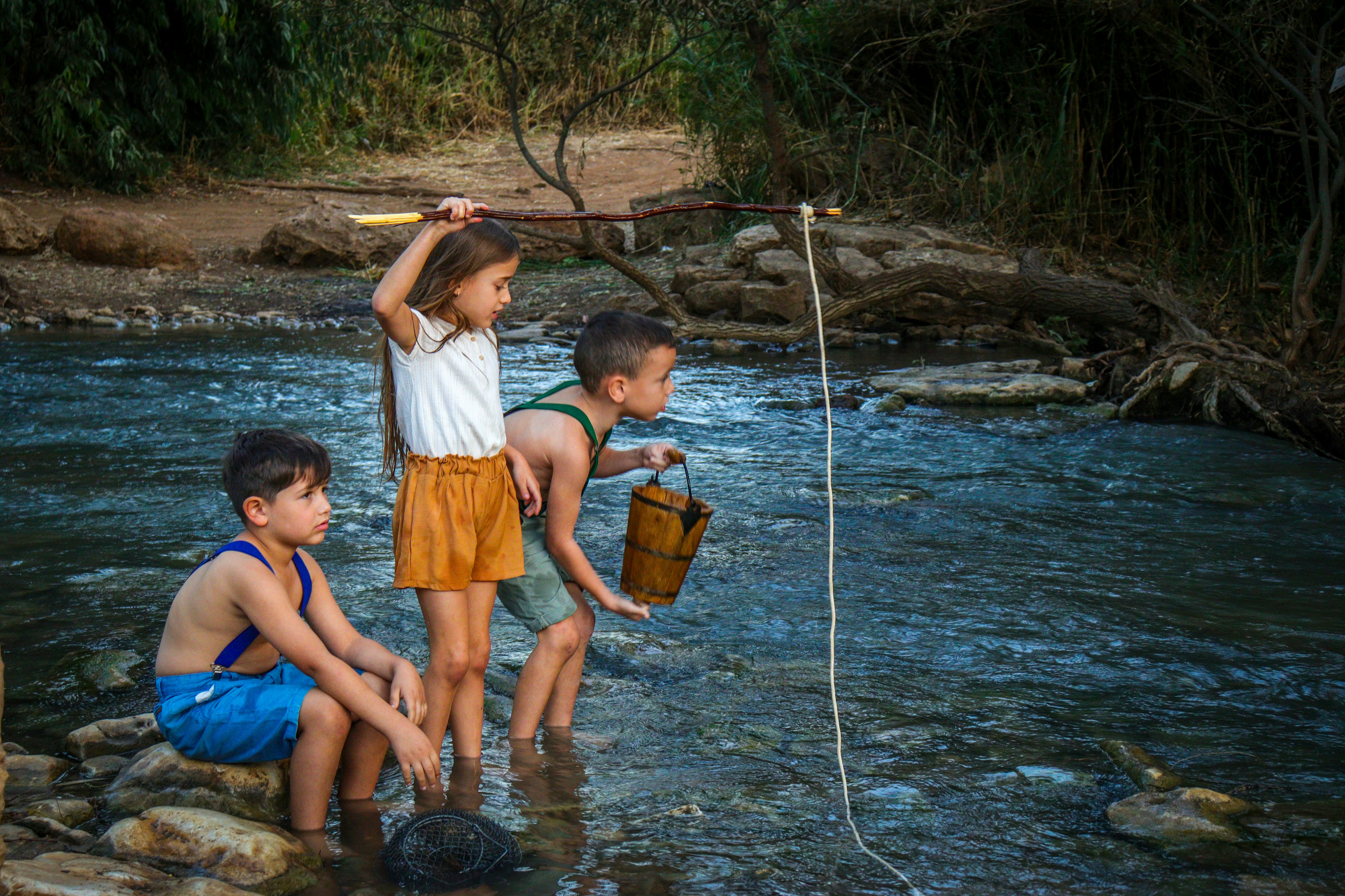 Three kids by a shallow river, one holding a rope and a bucket, with water and rocks visible in the foreground and trees in the background.