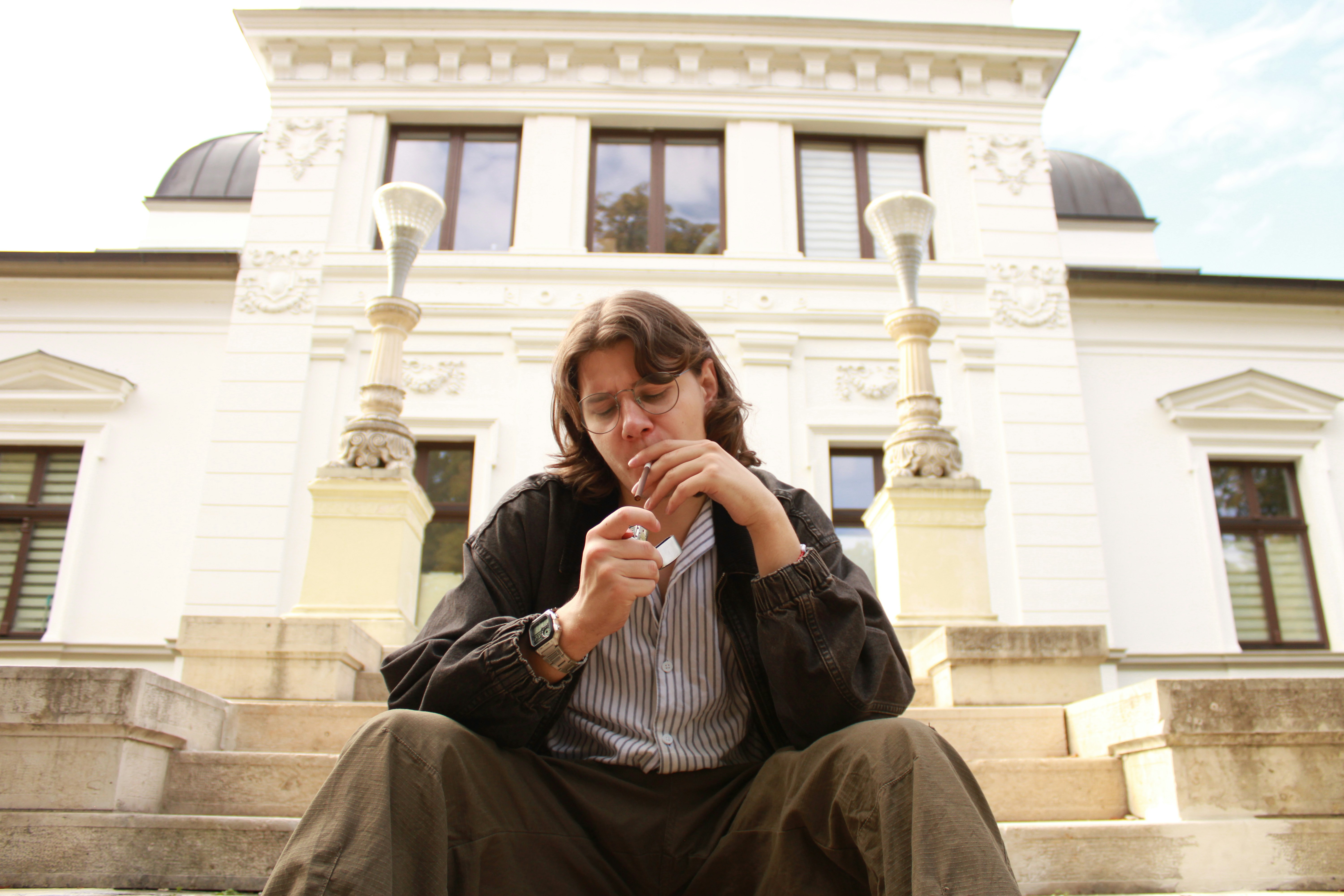 A man sits on grand stone steps in front of a white neoclassical building, lighting a cigarette with the camera at eye level, capturing a candid, relaxed moment.