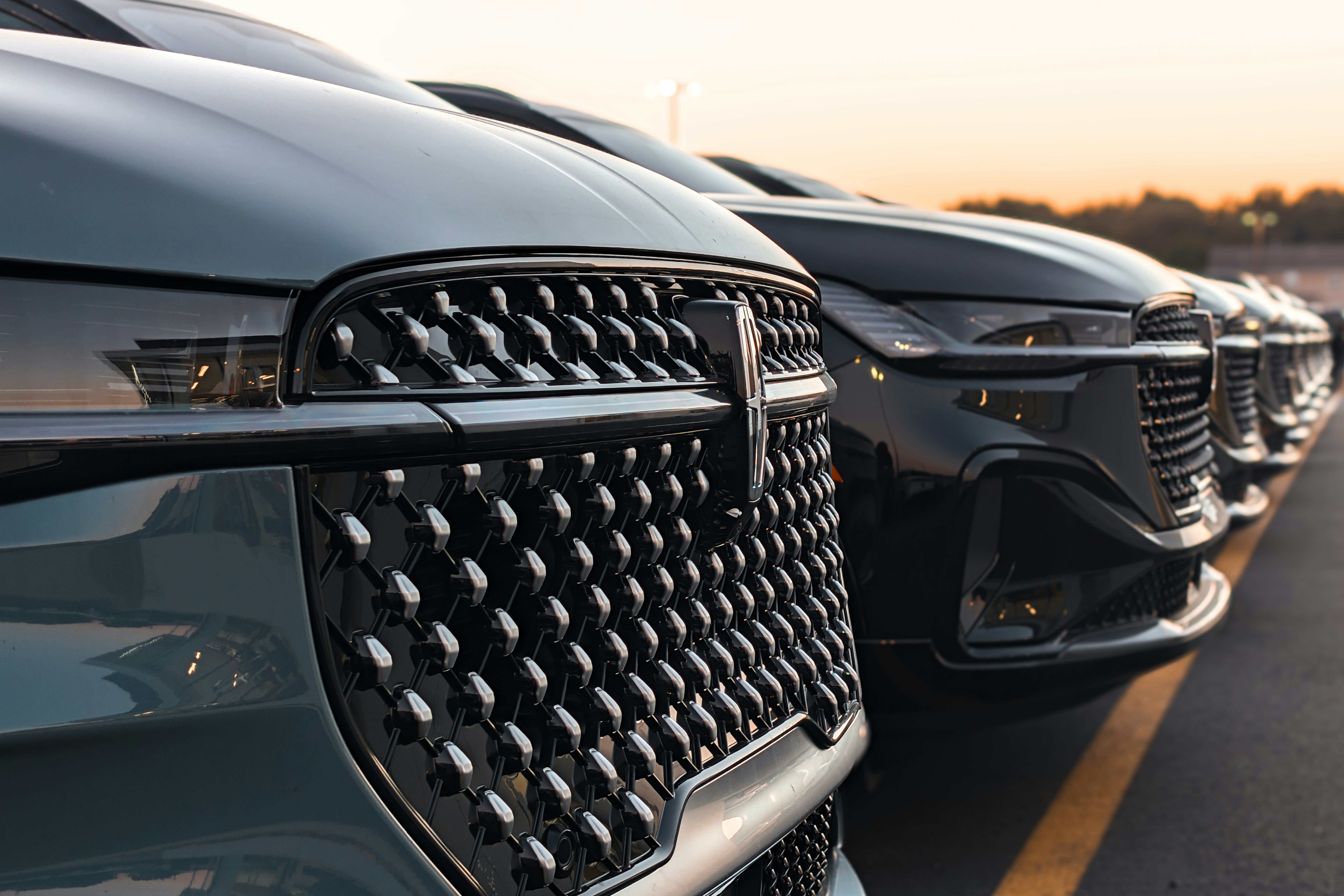 This macro photograph highlights the intricate metallic grille and vertical emblem of a sleek luxury SUV, leading an aligned fleet of vehicles across dealership asphalt during a warm vivid sunset.