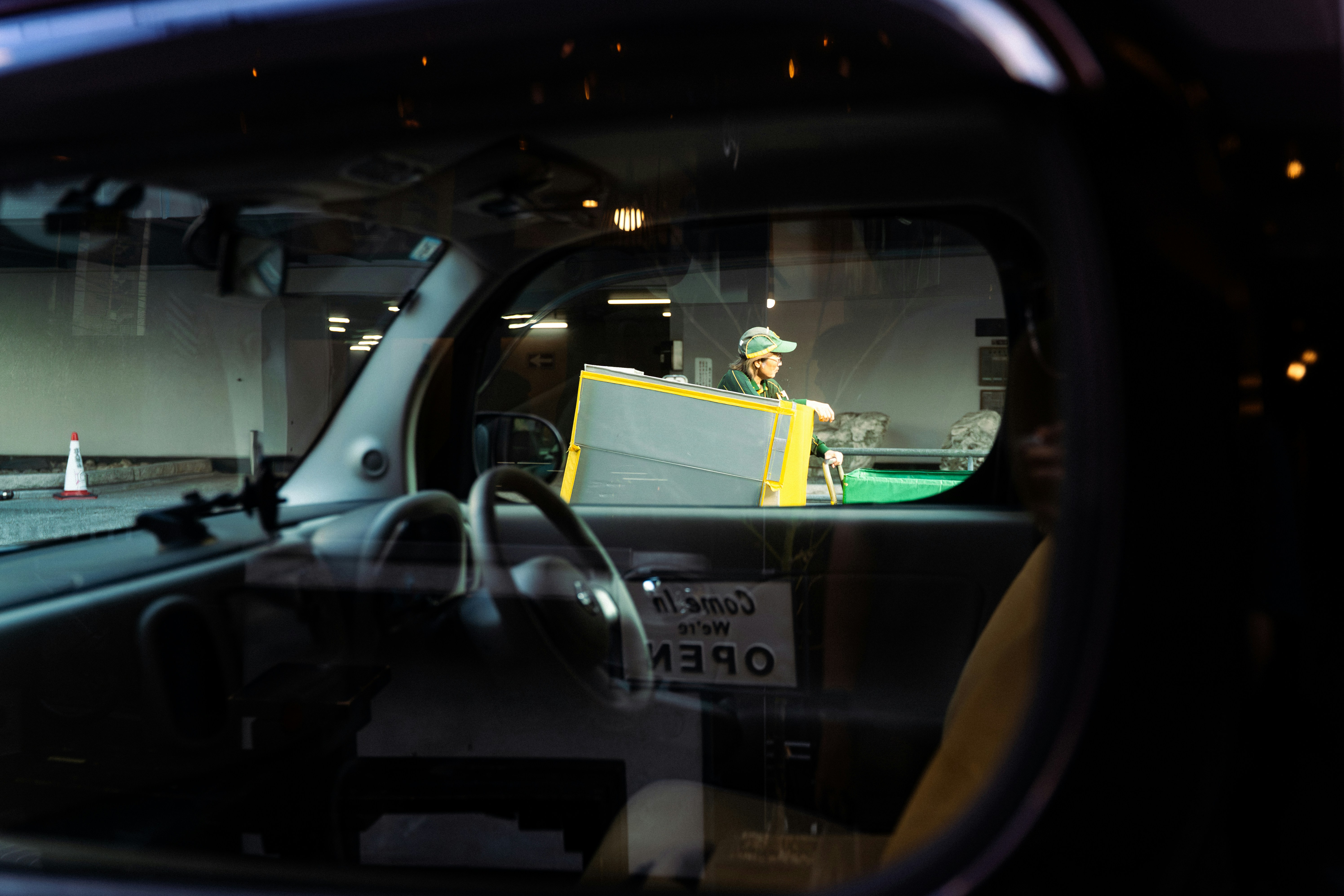 Holding a reflective moment inside a vehicle, a workspace with a person and equipment is visible through the front window; the scene captures a contrast between interior details and a bright, green-edged object outside. This is a photographic capture inside a showroom or garage.