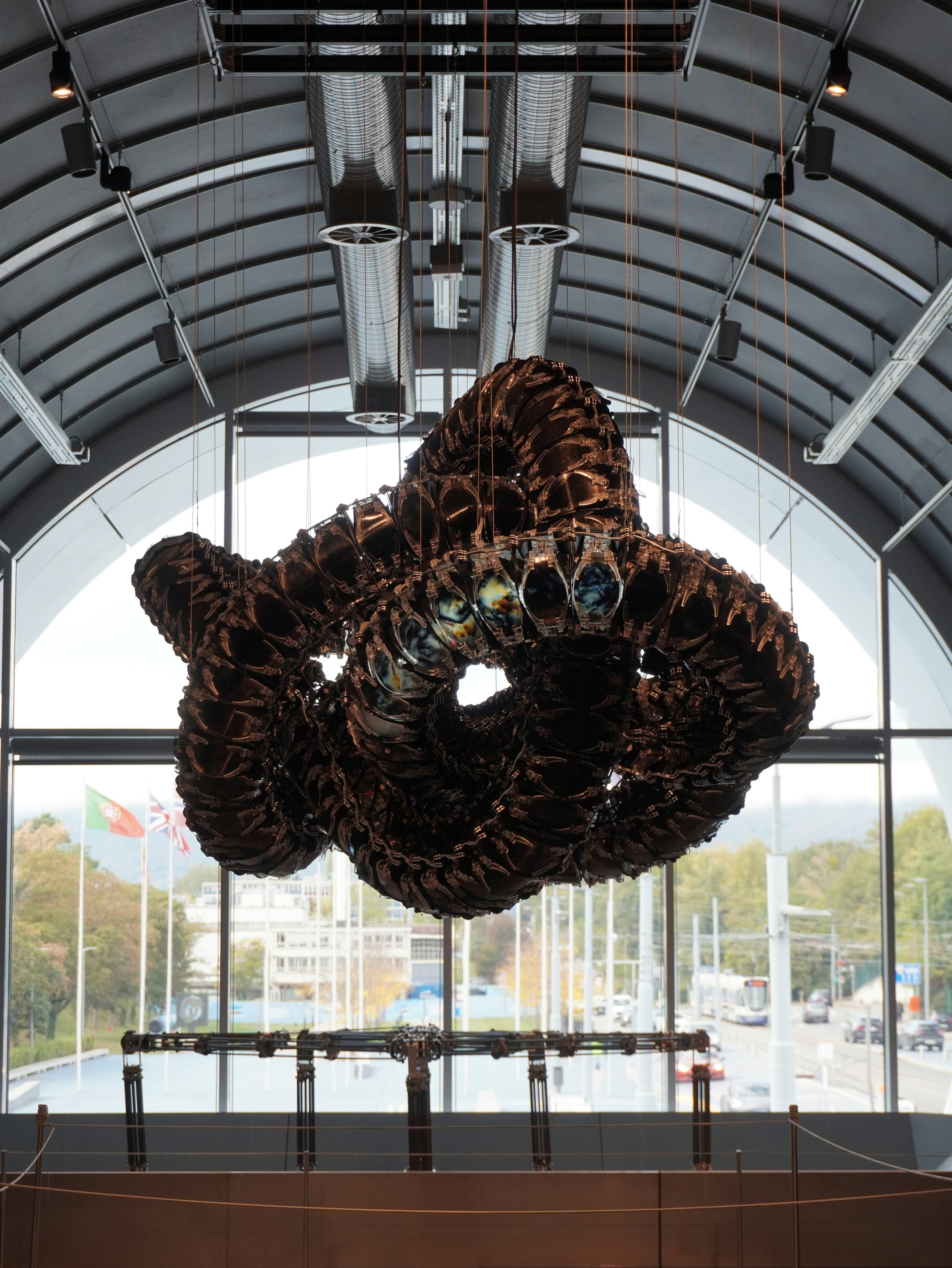 Dark, spiraling sculpture suspended in a bright atrium with glass walls and overhead industrial lighting. A modern art installation framed by large windows and flags outside; the piece hangs centrally above a railing.