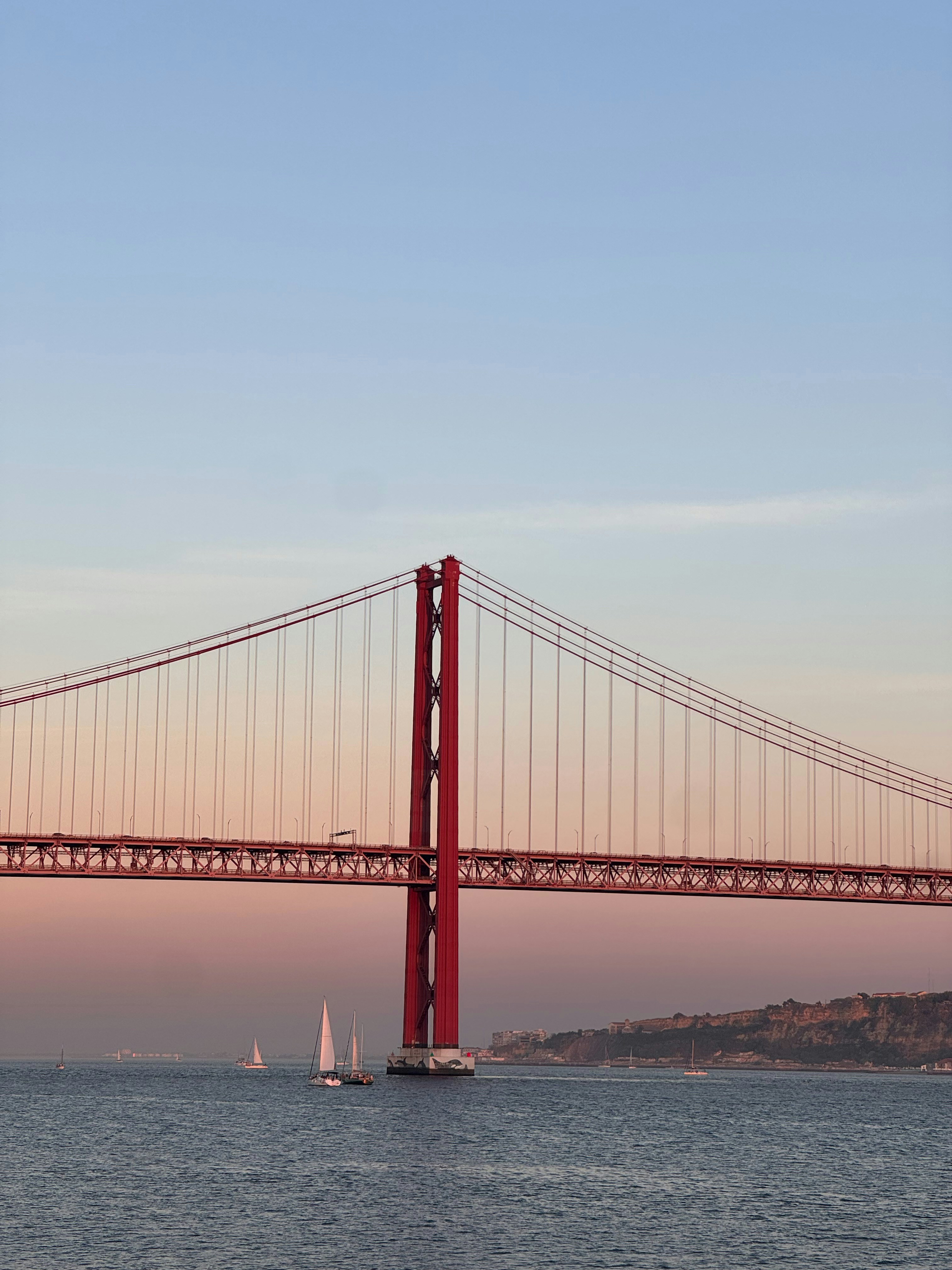 Suspension bridge spans calm harbor with sailboats nearby; pastel sky transitions from blue to warm pink as evening settles over the water.