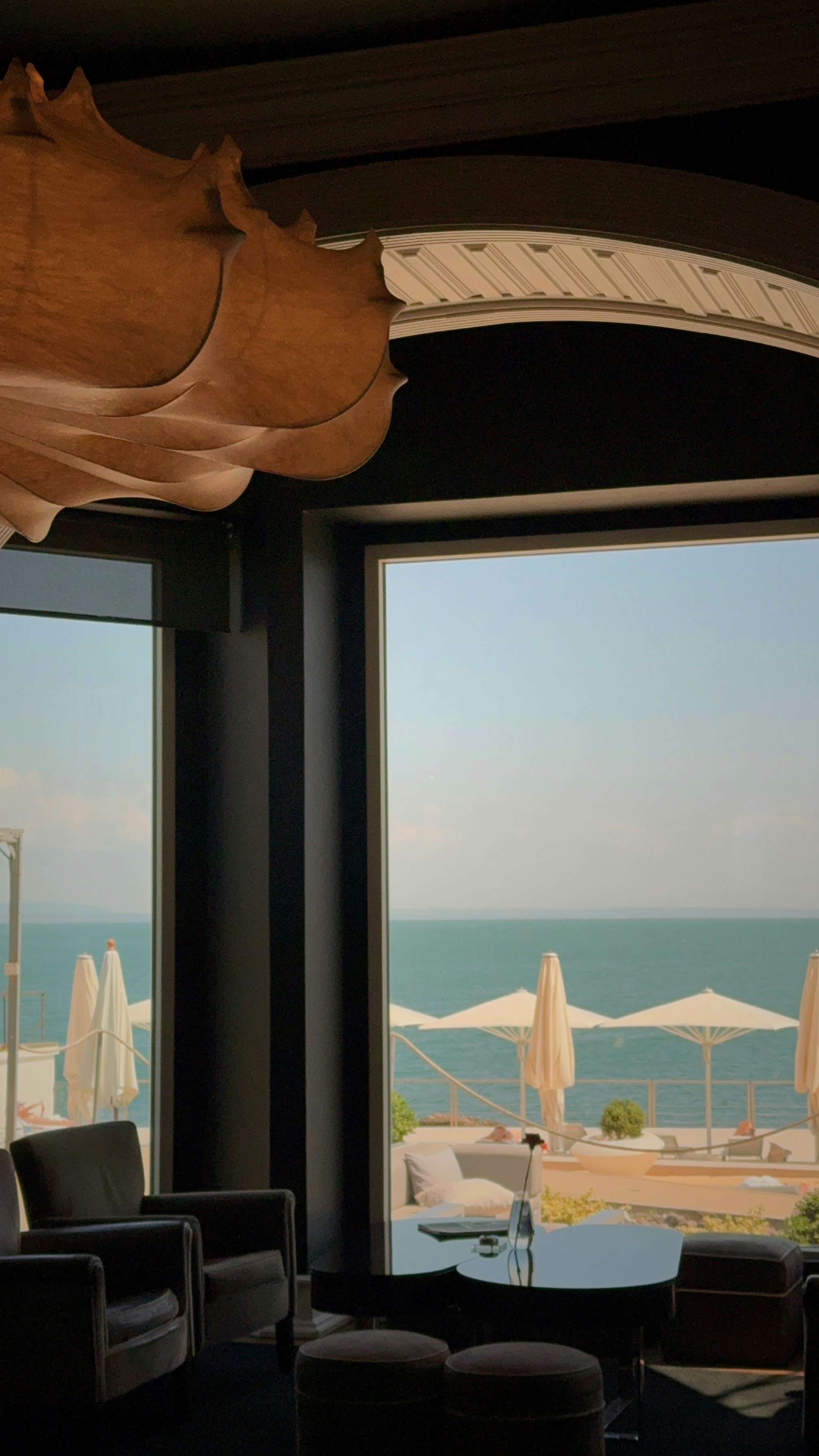 Dark interior lounge with a large window opening to a bright seascape and umbrellas on the beach beyond.