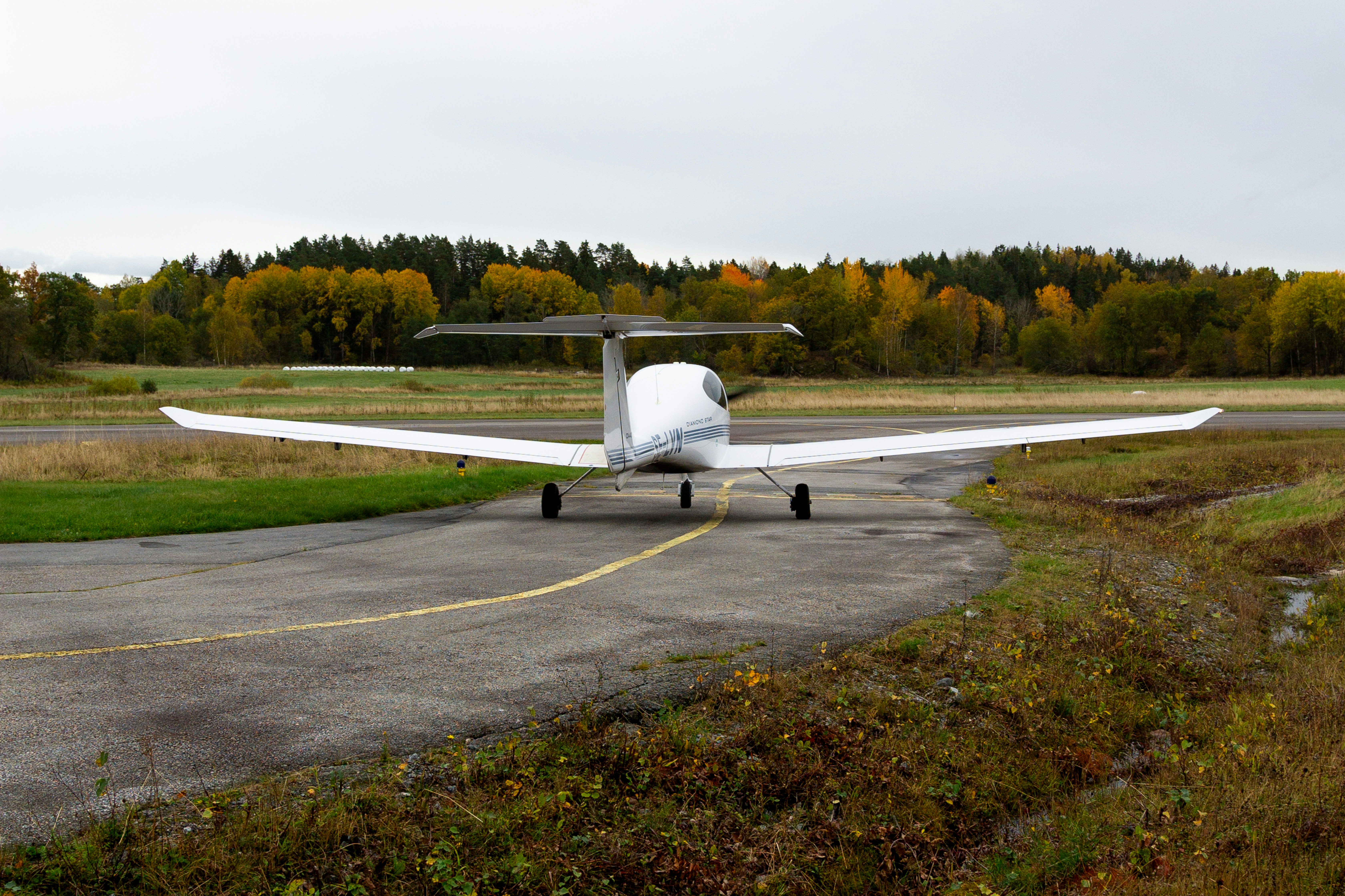 White glider sits on a cracked asphalt runway with yellow markings, set against a backdrop of autumn trees under an overcast sky.
