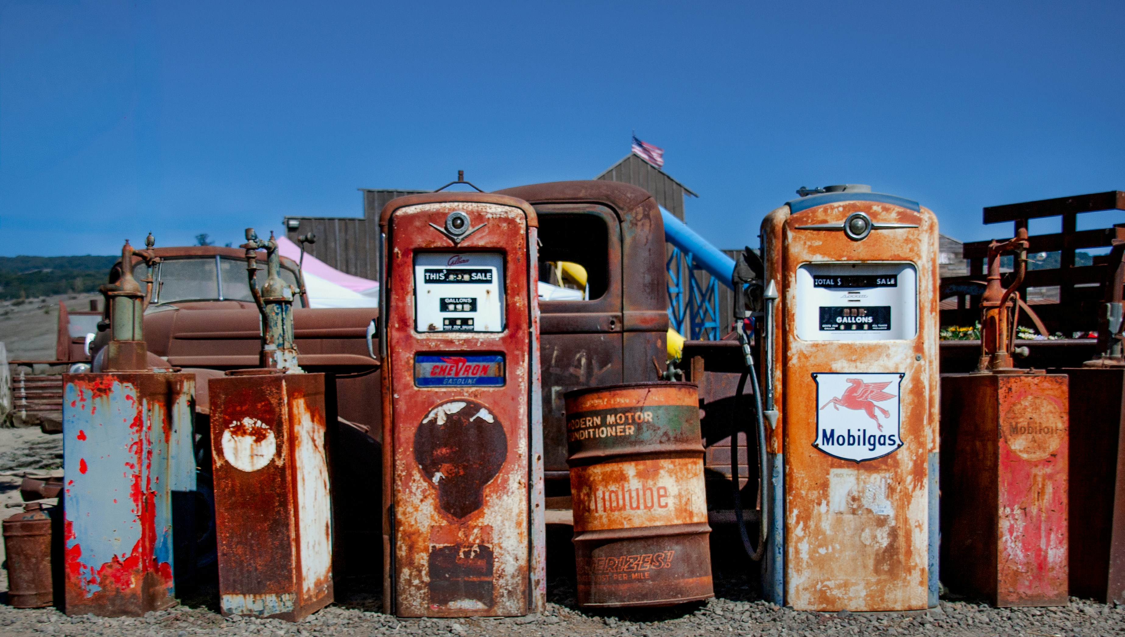 Row of rusty vintage gas pumps and barrels at a desert-mawn roadside station, with weathered paint and a faded Mobilgas label; a retro fuel-station scene.