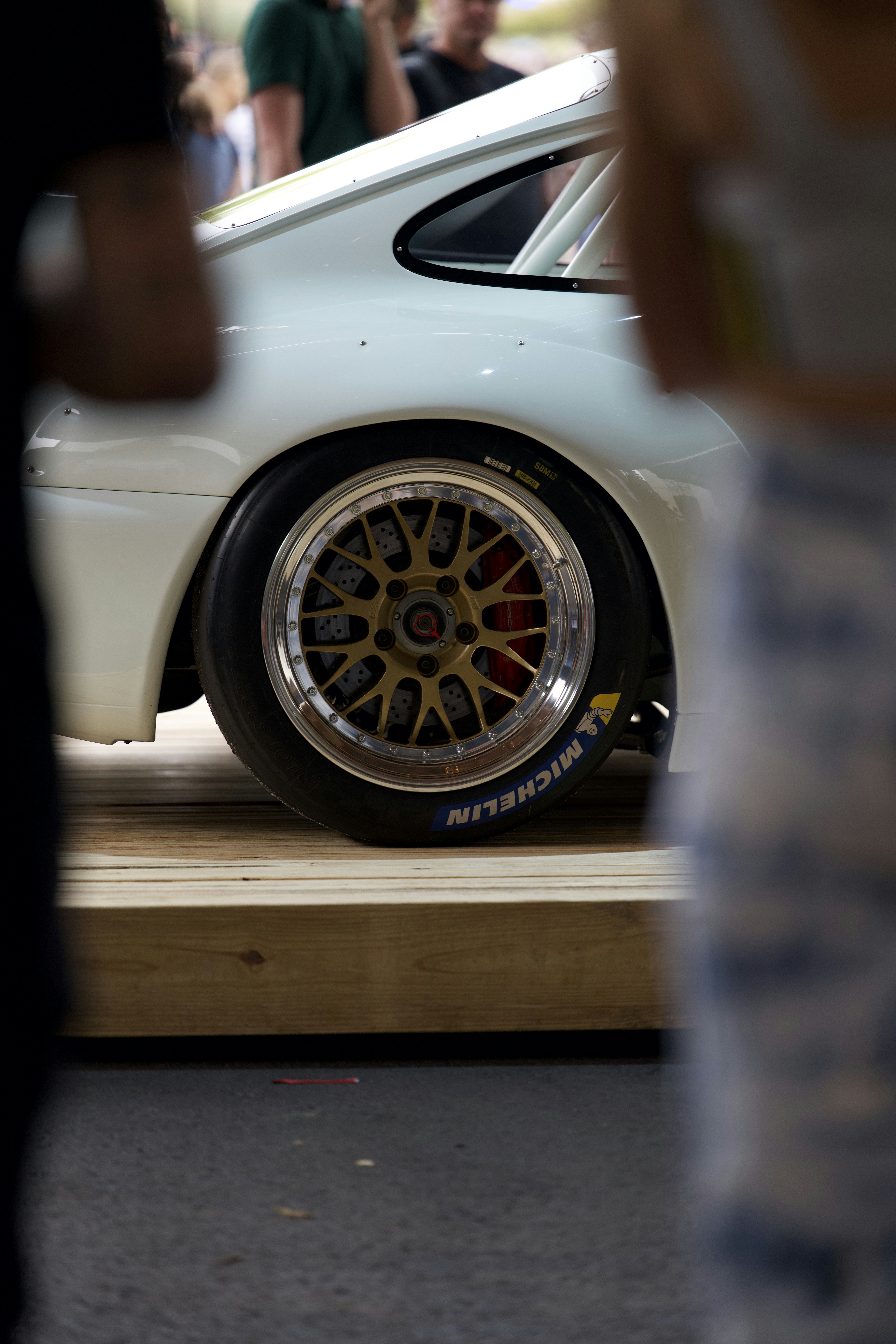 Close-up of a sports car wheel with gold spokes and a red brake caliper, framed between passing crowds; a shallow depth of field highlights the vehicle while the surrounding spectators blur into the background.
