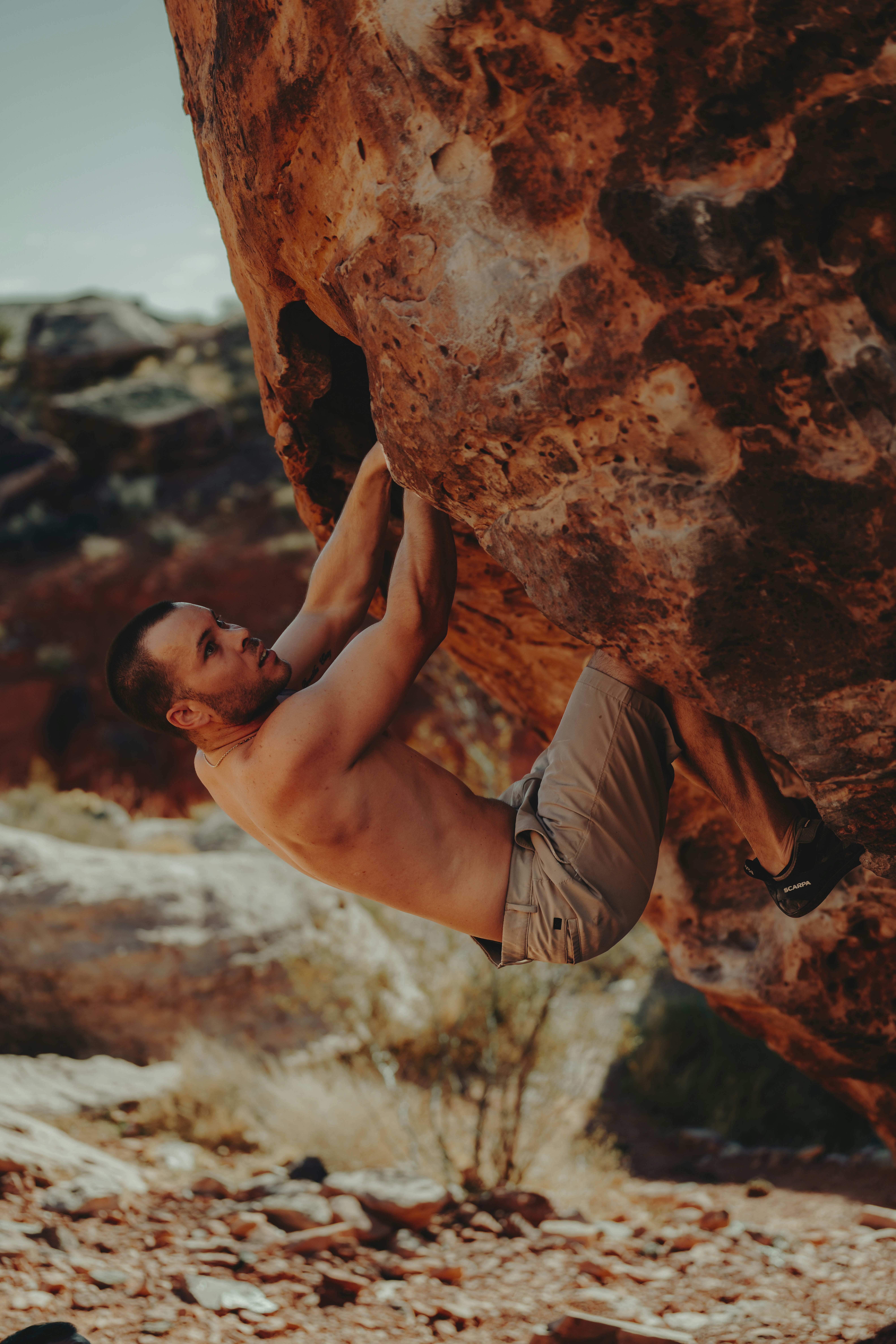 A shirtless man bouldering on a large red sandstone rock in a desert landscape
