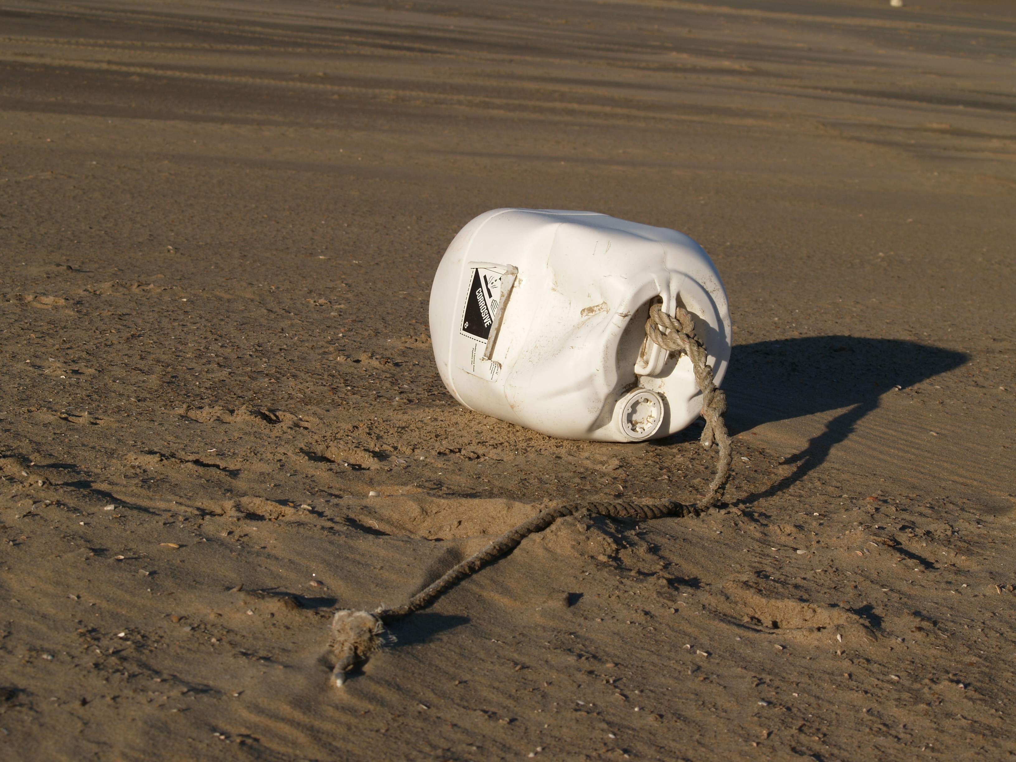 A white plastic container lies half-buried on a sunlit beach, tethered by a frayed rope that trails across the sand.