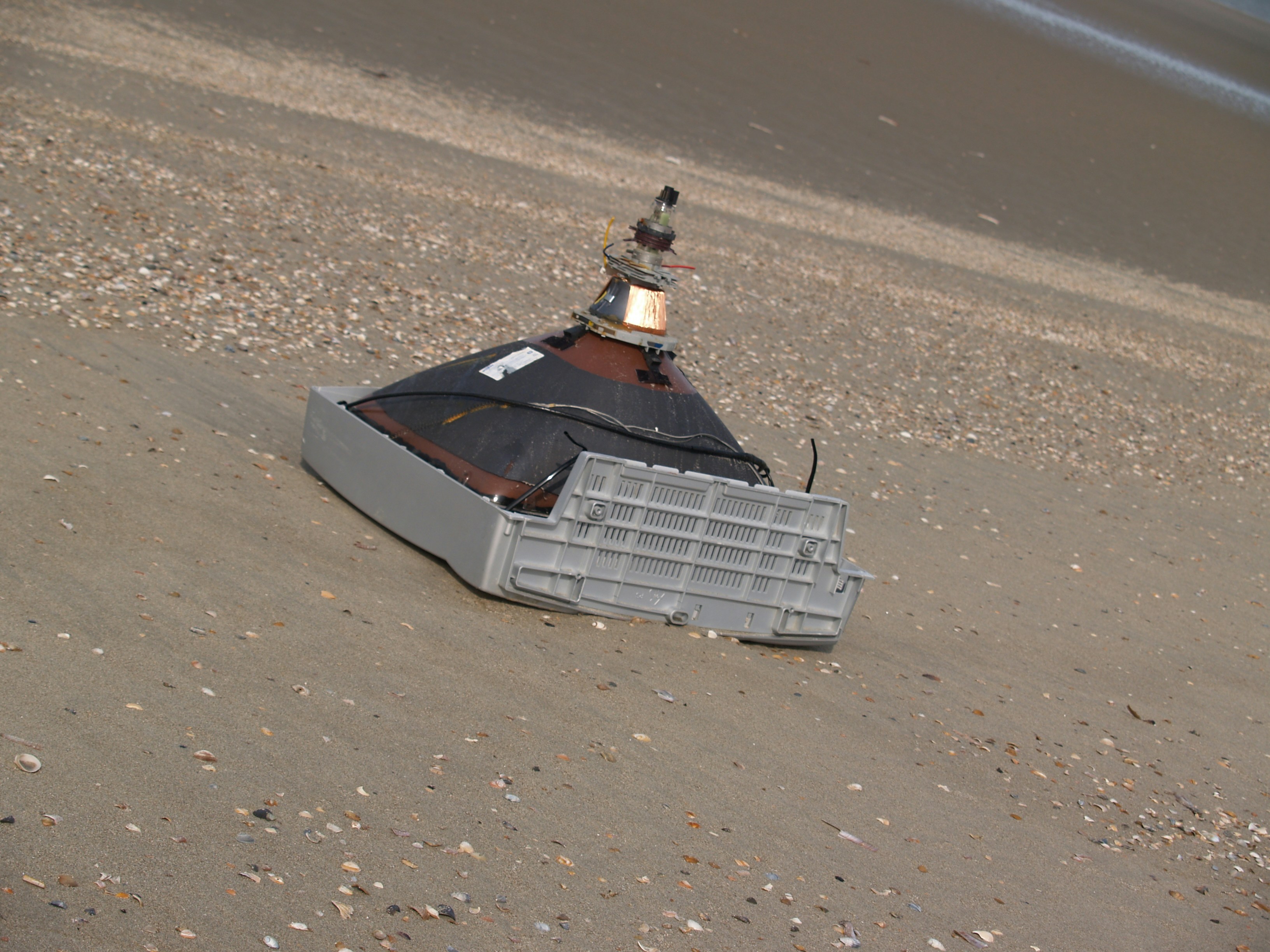A weathered buoy-lamp lies stranded on a sandy beach, its ribs and casing relaxed in the open air. The scene emphasizes abandonment and the quiet rhythm of shoreline debris.