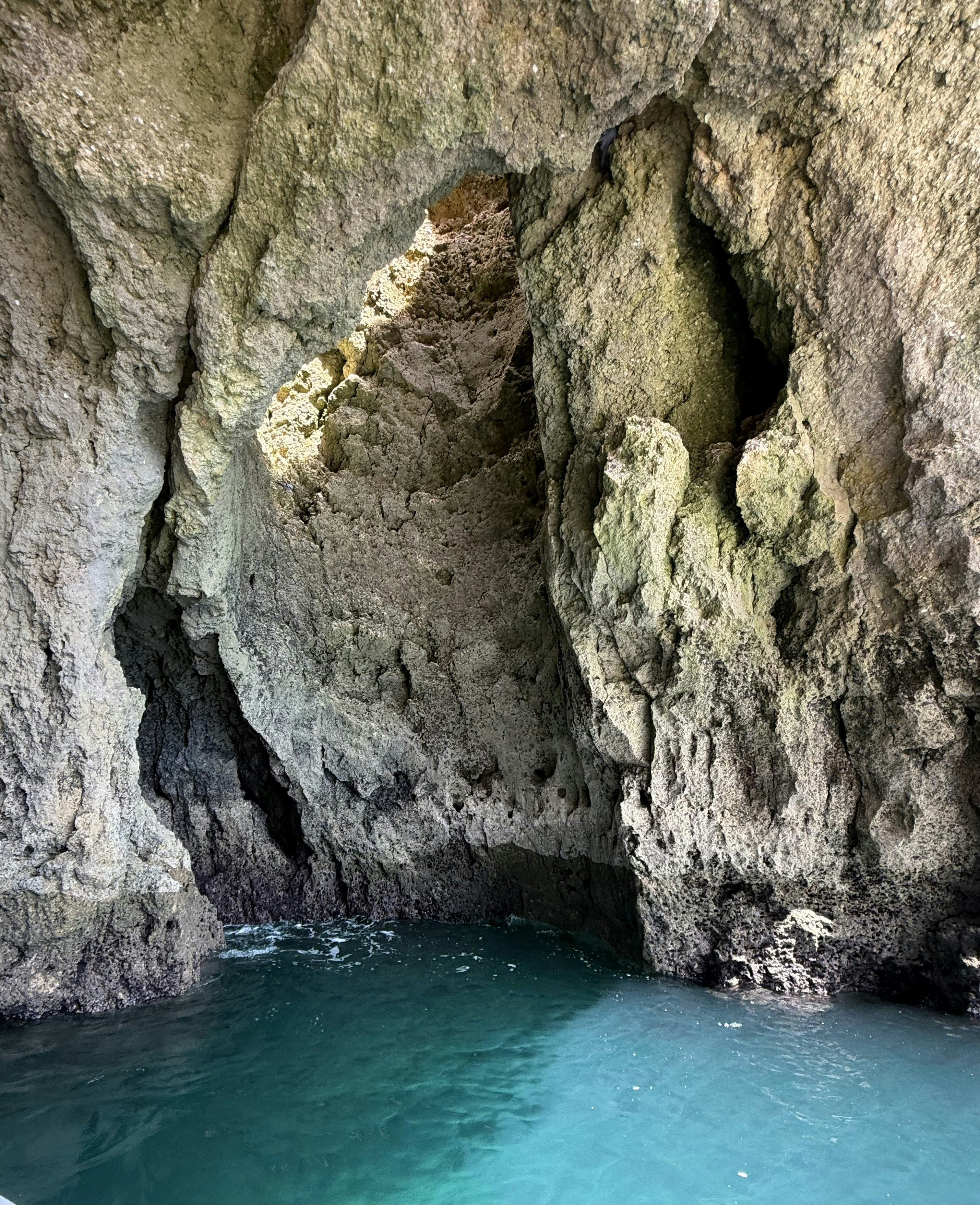Turquoise water laps at the base of rugged limestone arches and a natural sea cave, sunlight illuminating textured rock faces.