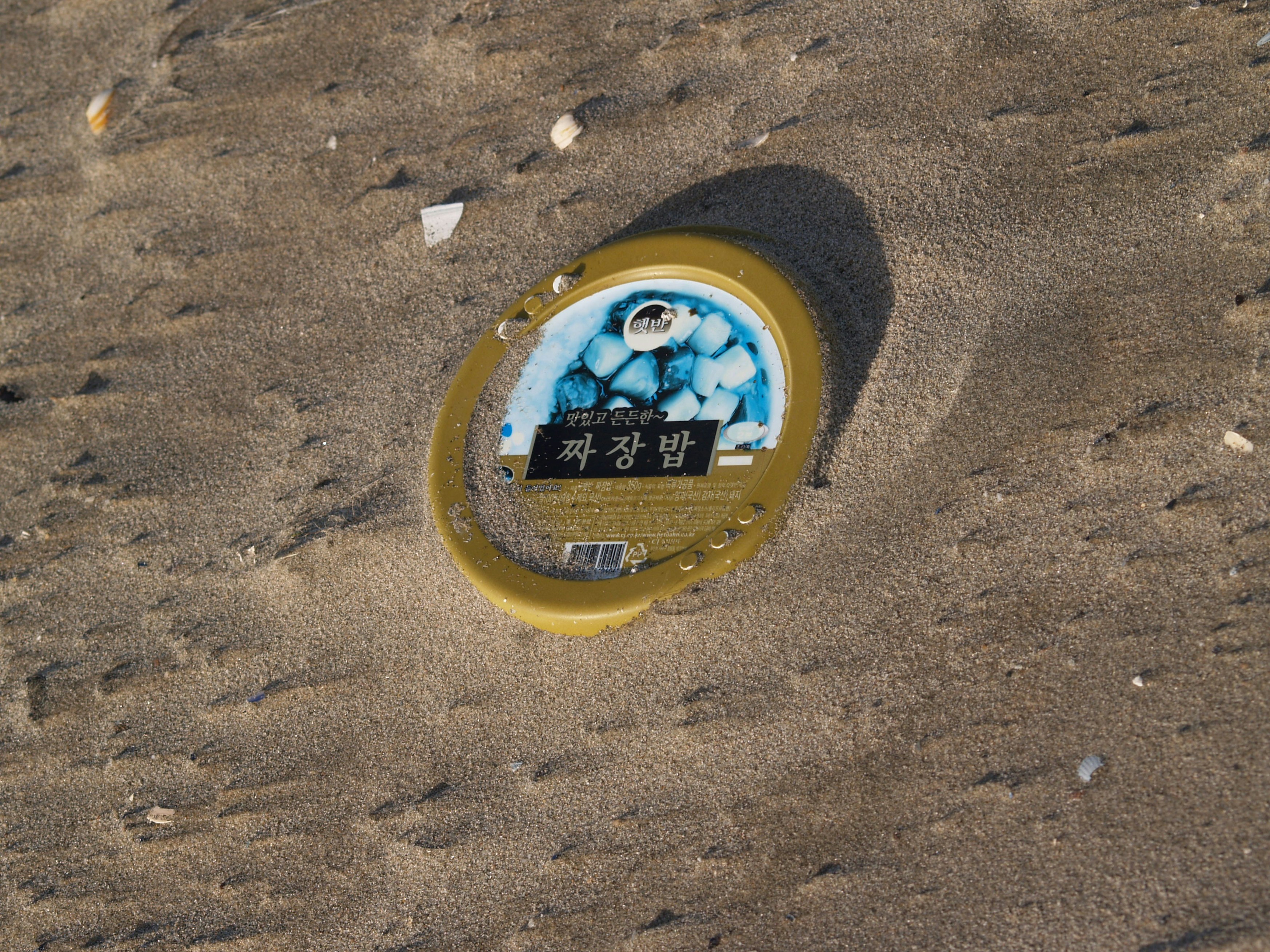A circular yellow-lidded container lies partially buried in sunlit sand, with a blue label visible inside. The scene emphasizes litter disposal in a coastal environment.