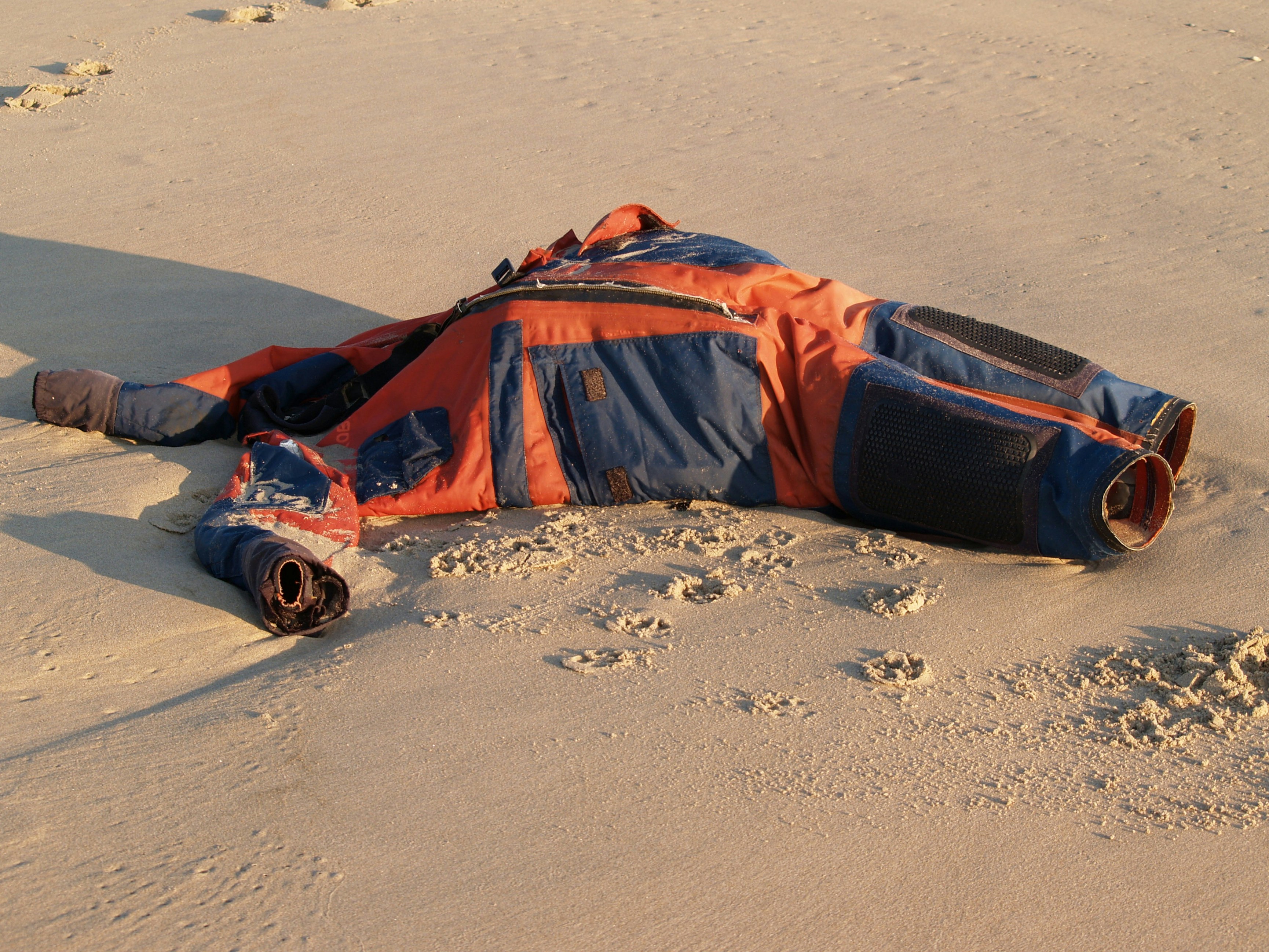 Orange and blue outdoor gear lies abandoned in fine sand, footprints trailing toward the horizon under warm late-afternoon light.