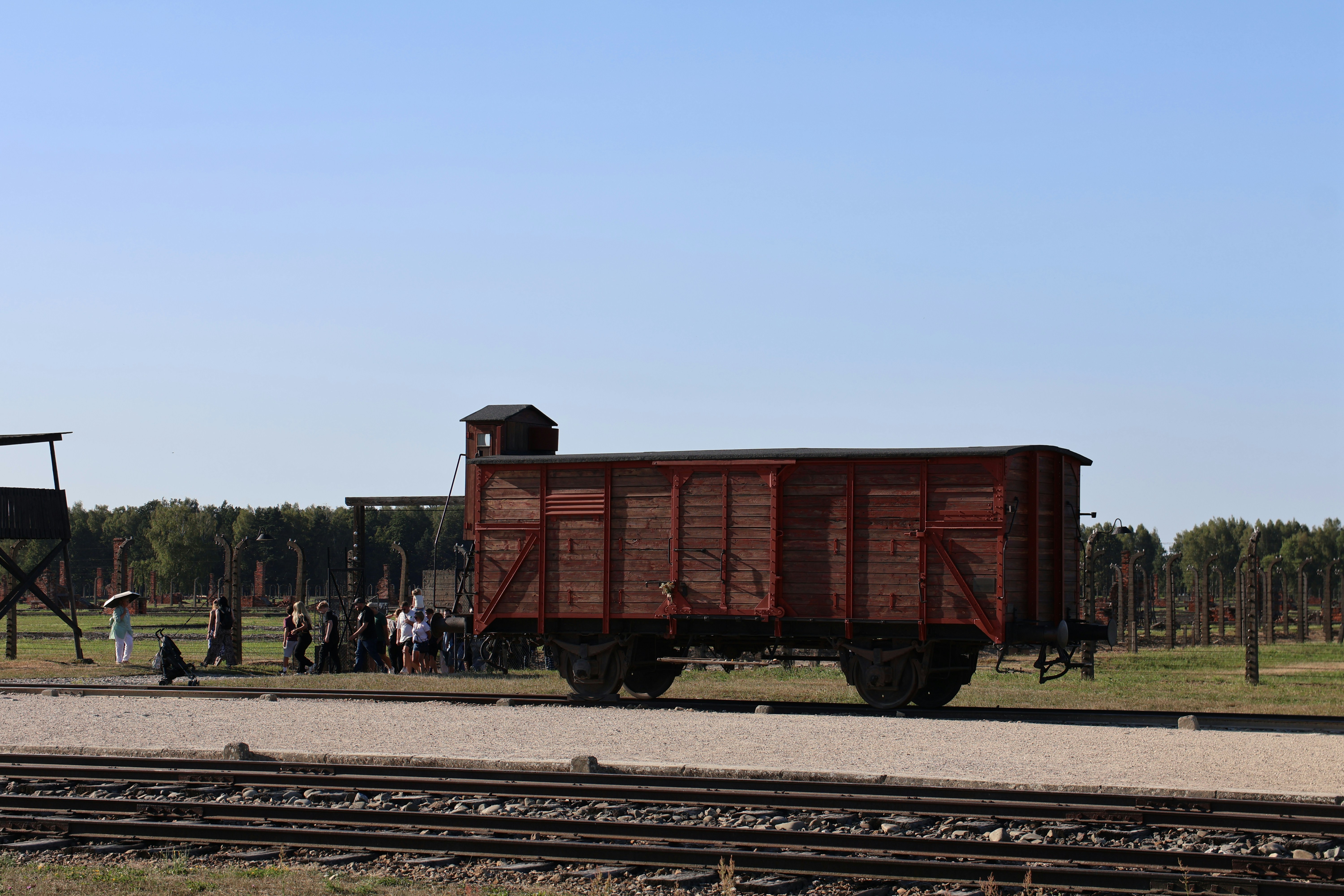 Rusty freight car on tracks with a distant group of people and a watchtower under a clear sky. A documentary scene of rail yard life with muted colors and wide horizon.
