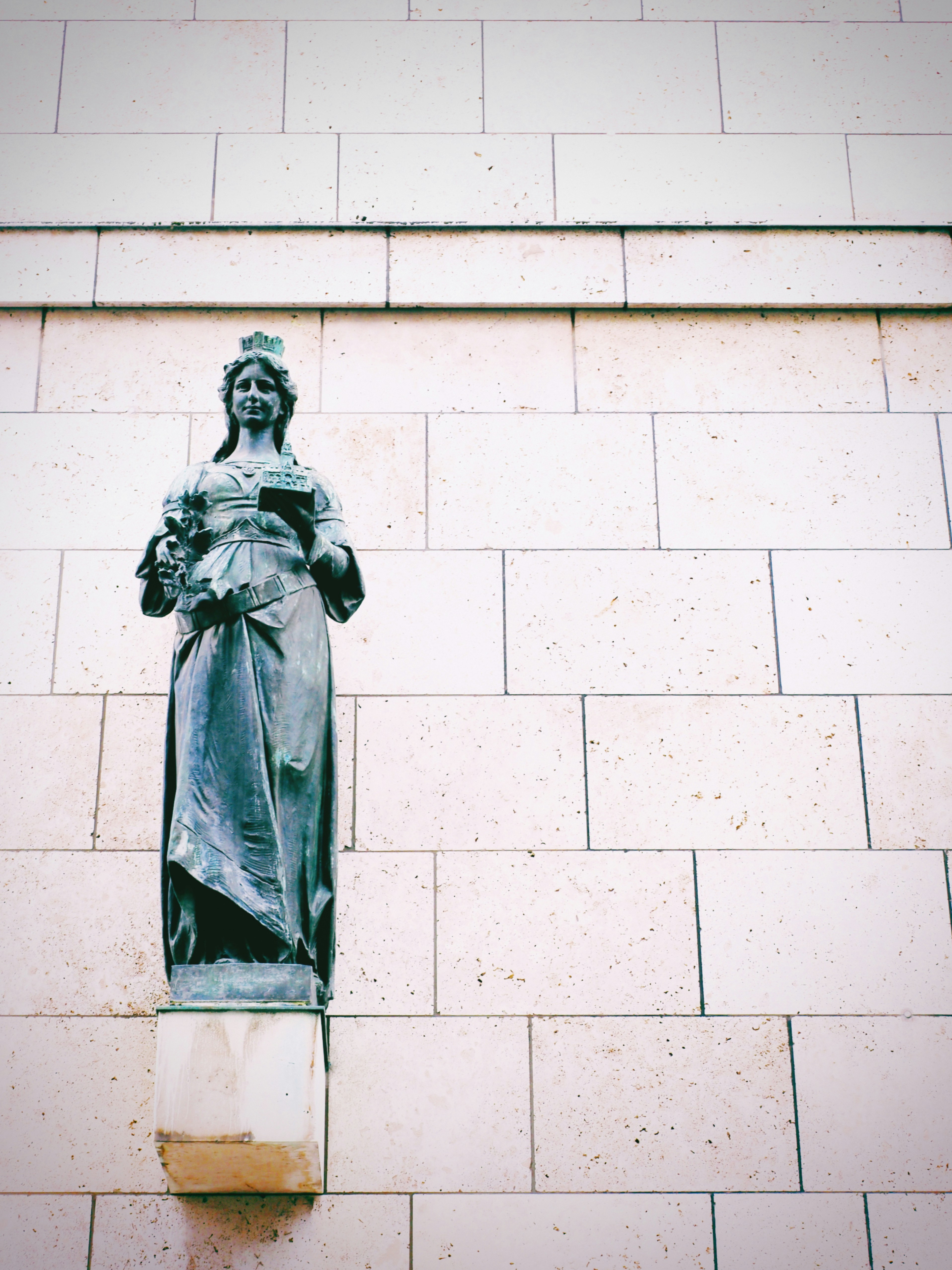 Statue of a crowned female figure holding an object, mounted on a stone pedestal against a tiled wall; the scene focuses on the sculpture with subtle color desaturation and soft contrast. This is a sculpture photograph.