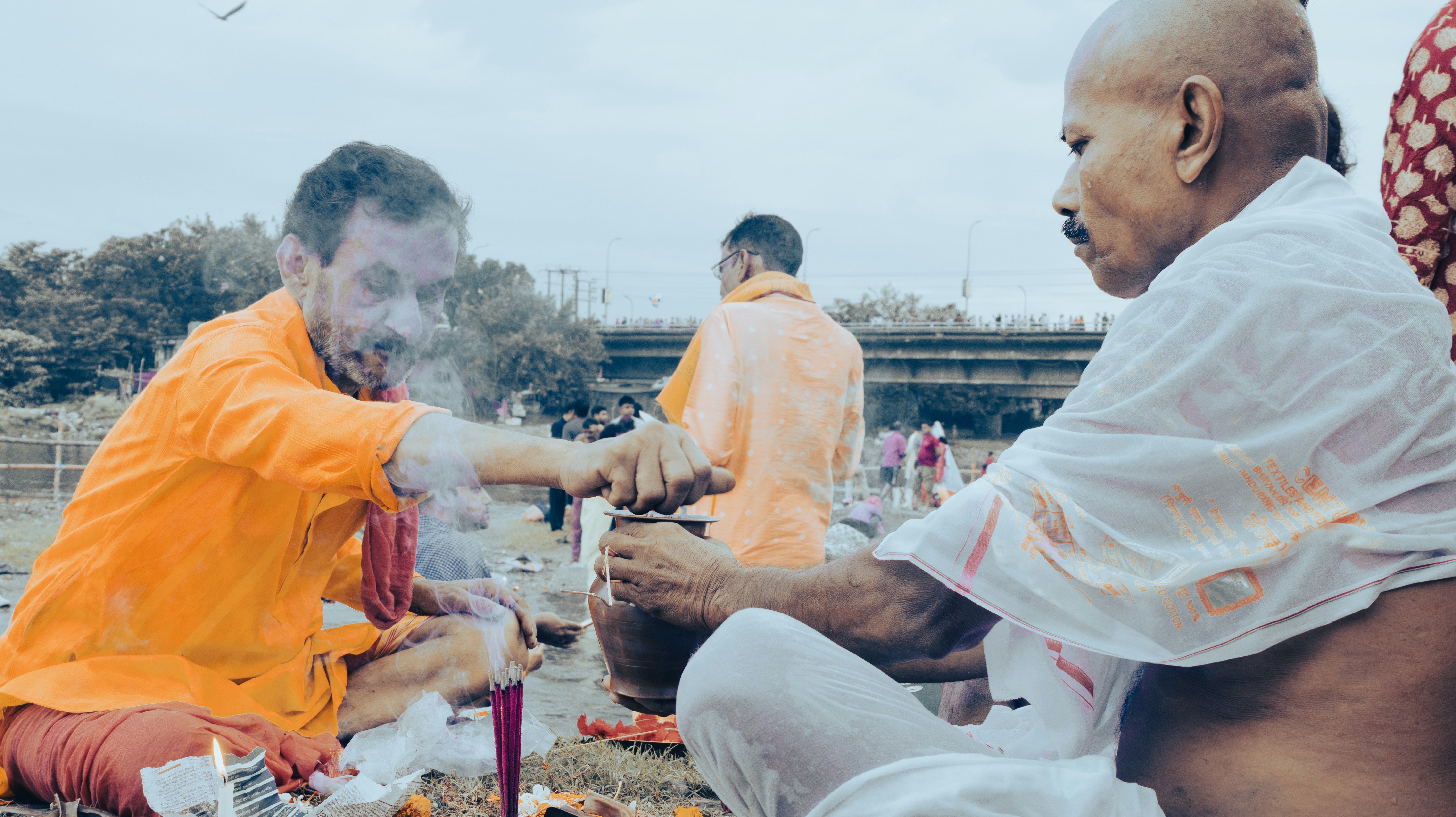 Two men sit cross-legged in a vibrant outdoor ritual, one in orange and the other in white, offering a ceremonial hand gesture with smoke and ritual vessels in the foreground.