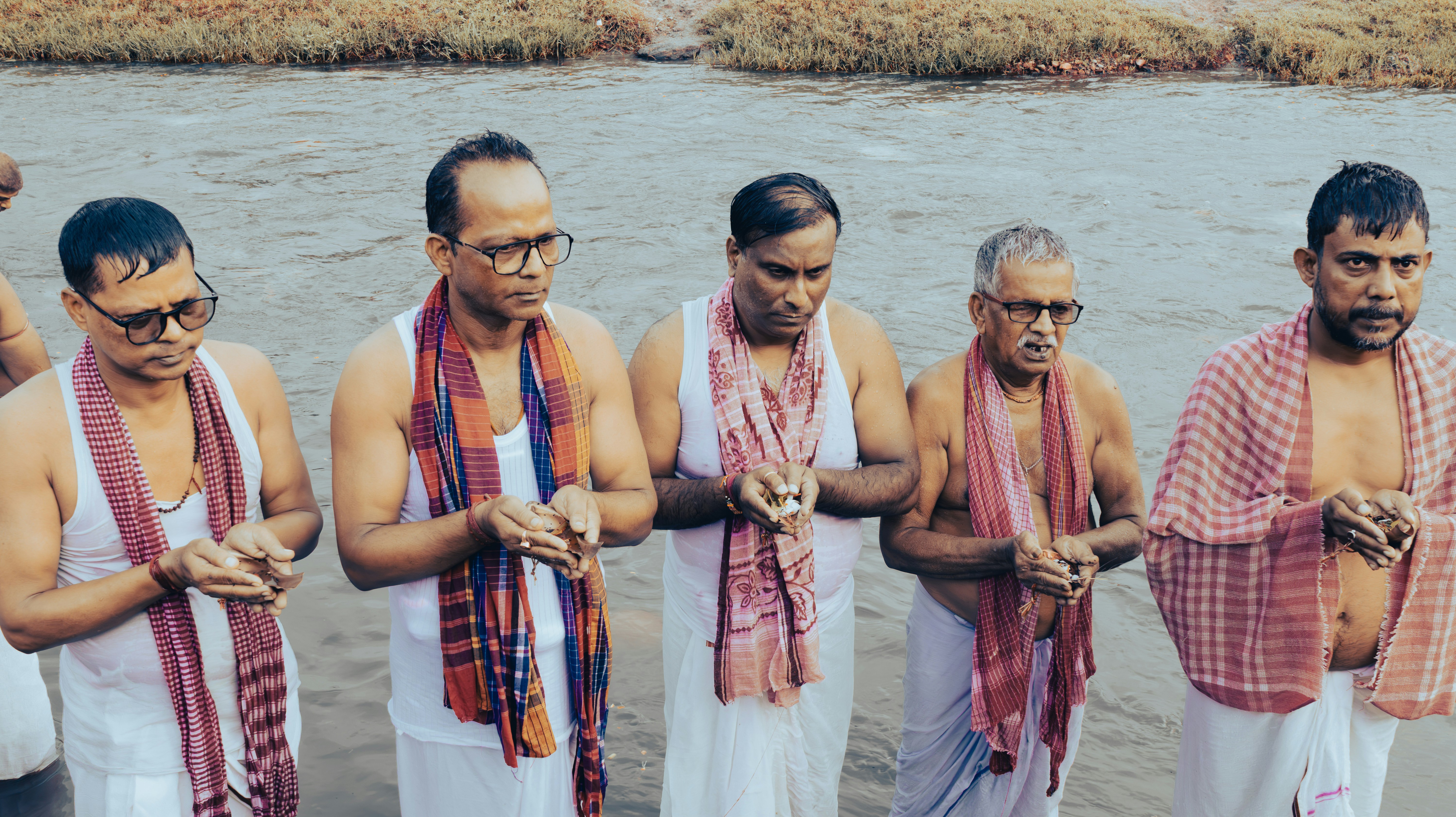 Five men in white robes with colorful shawls stand along a riverbank, hands clasped in prayer. The scene captures a communal ritual by the water.