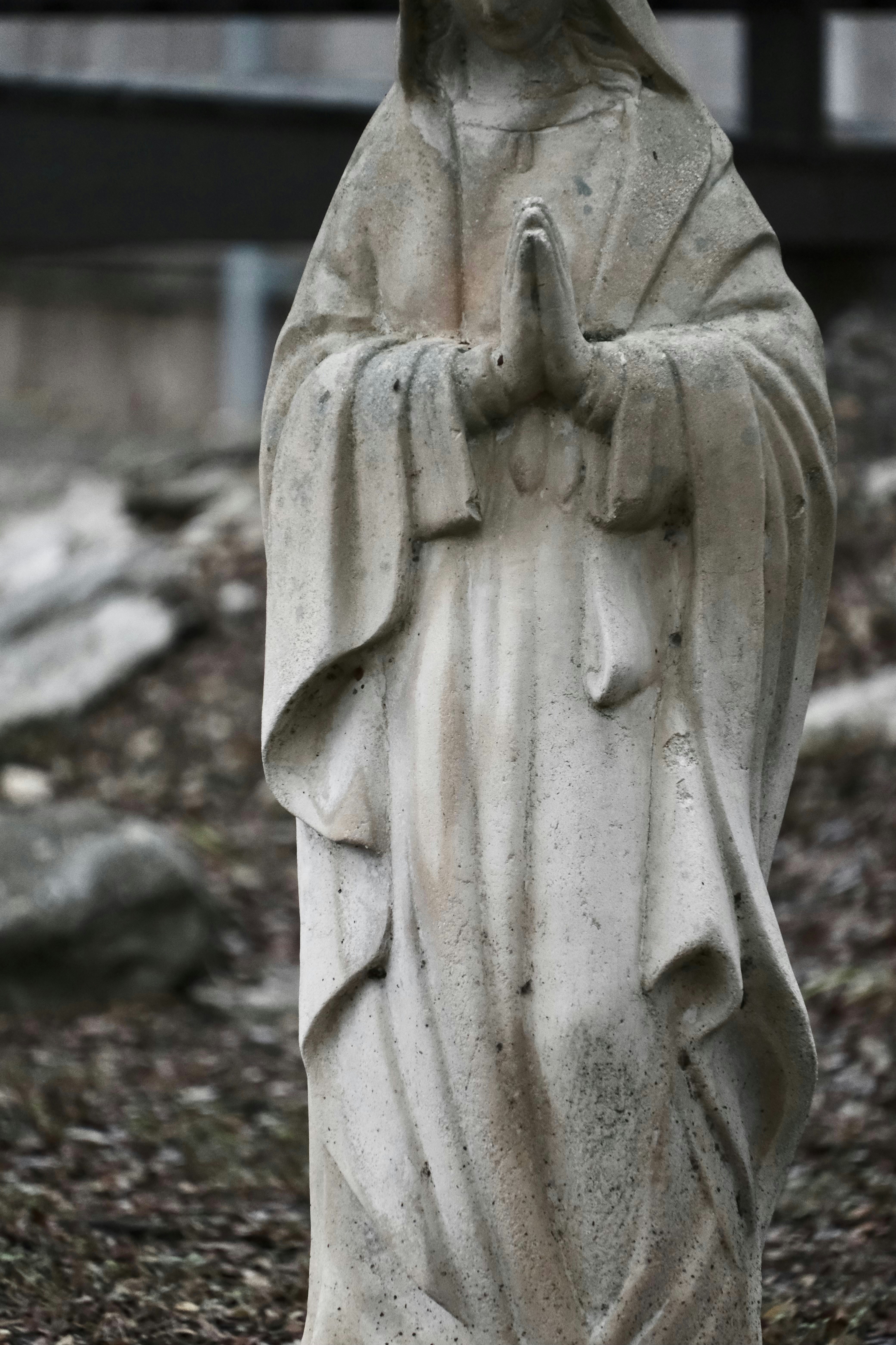 Weathered statue with hands in prayer stands in a muted garden setting, focused on the serene figure and its draped robes.