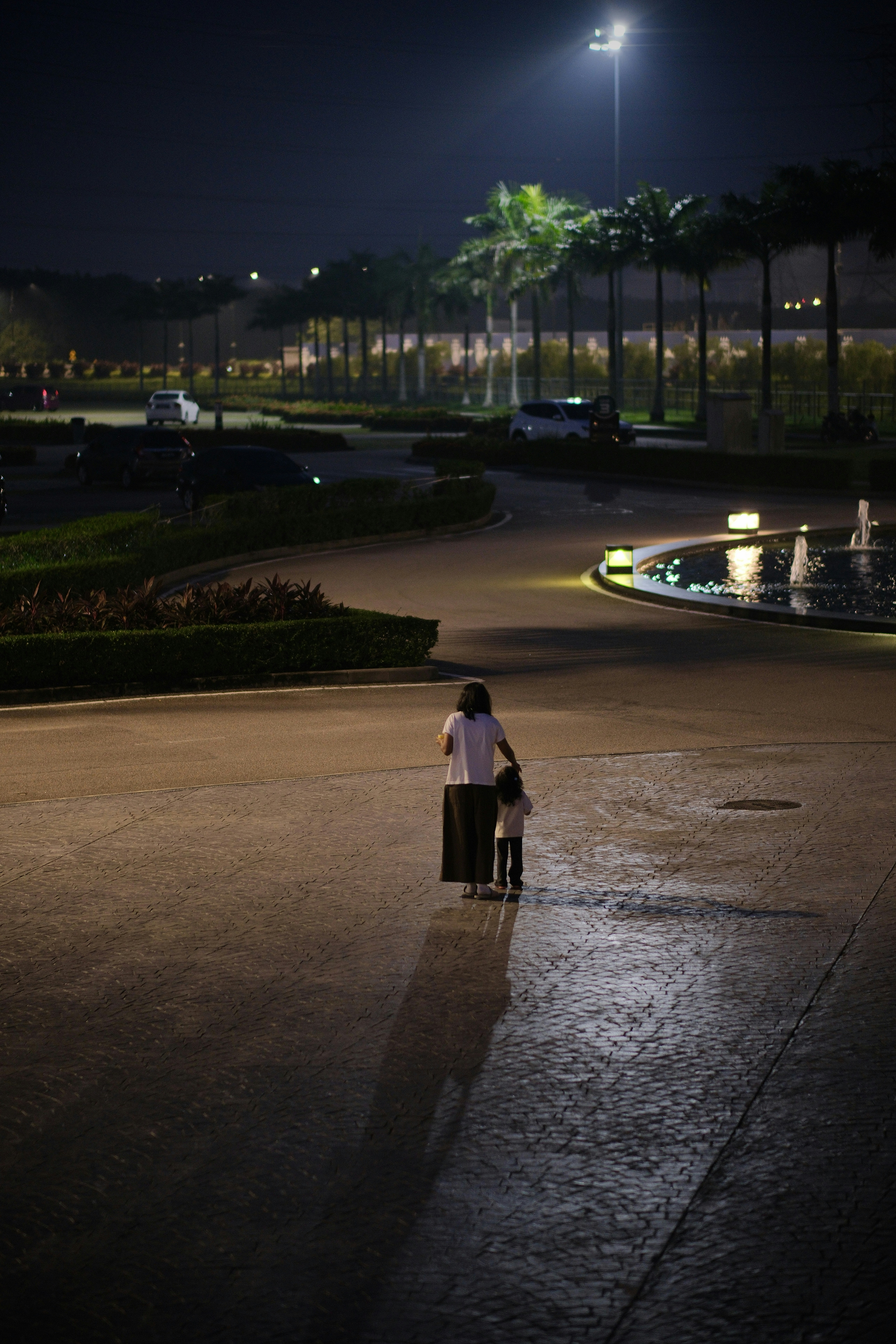 A mother and child stand together near a circular fountain at night, illuminated by ambient streetlights in a calm, urban plaza.