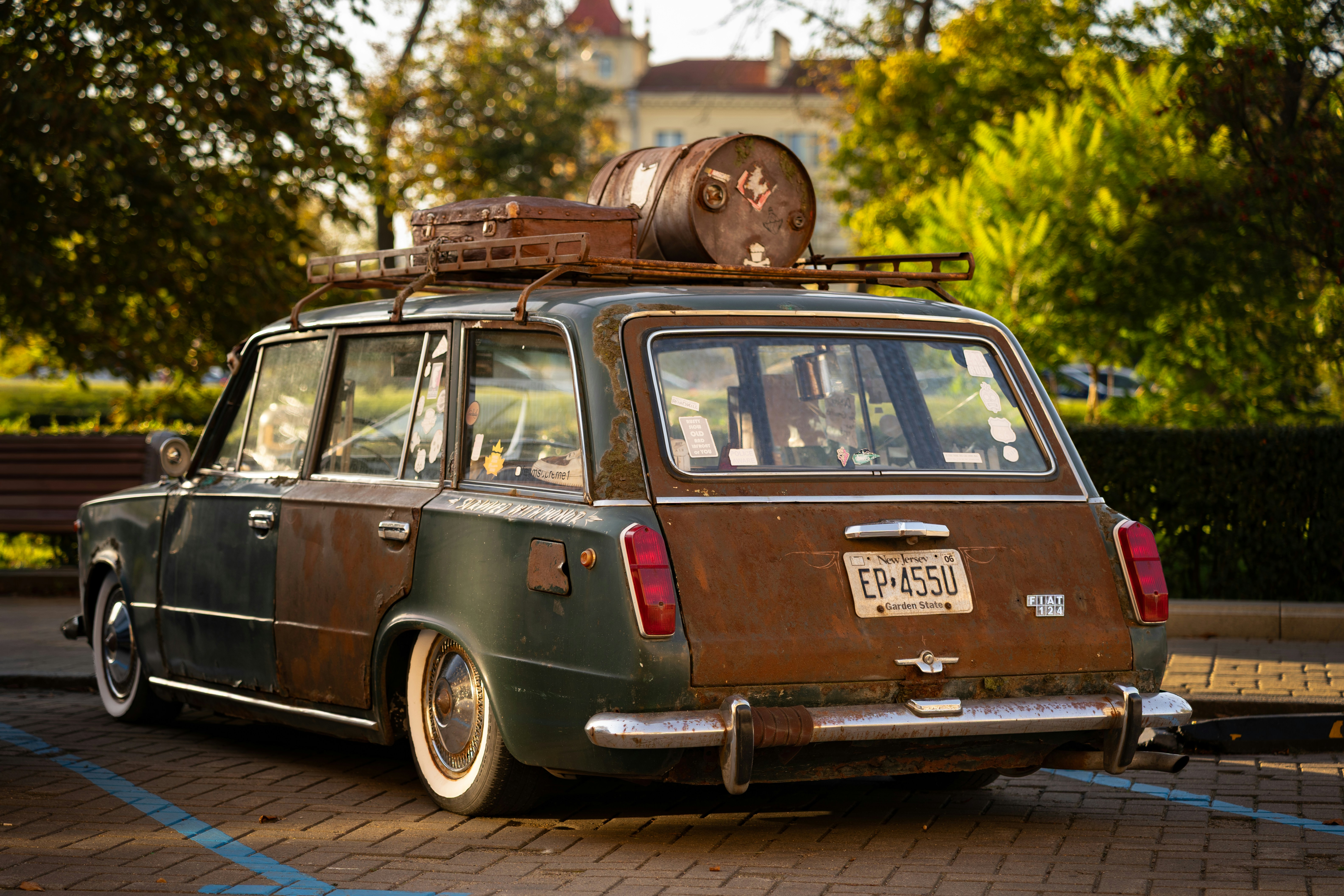 A weathered vintage station wagon sits parked in a lot, laden with luggage on its roof and a rusty spare wheel visible above. The scene emphasizes age, patina, and a quiet, nostalgic mood.