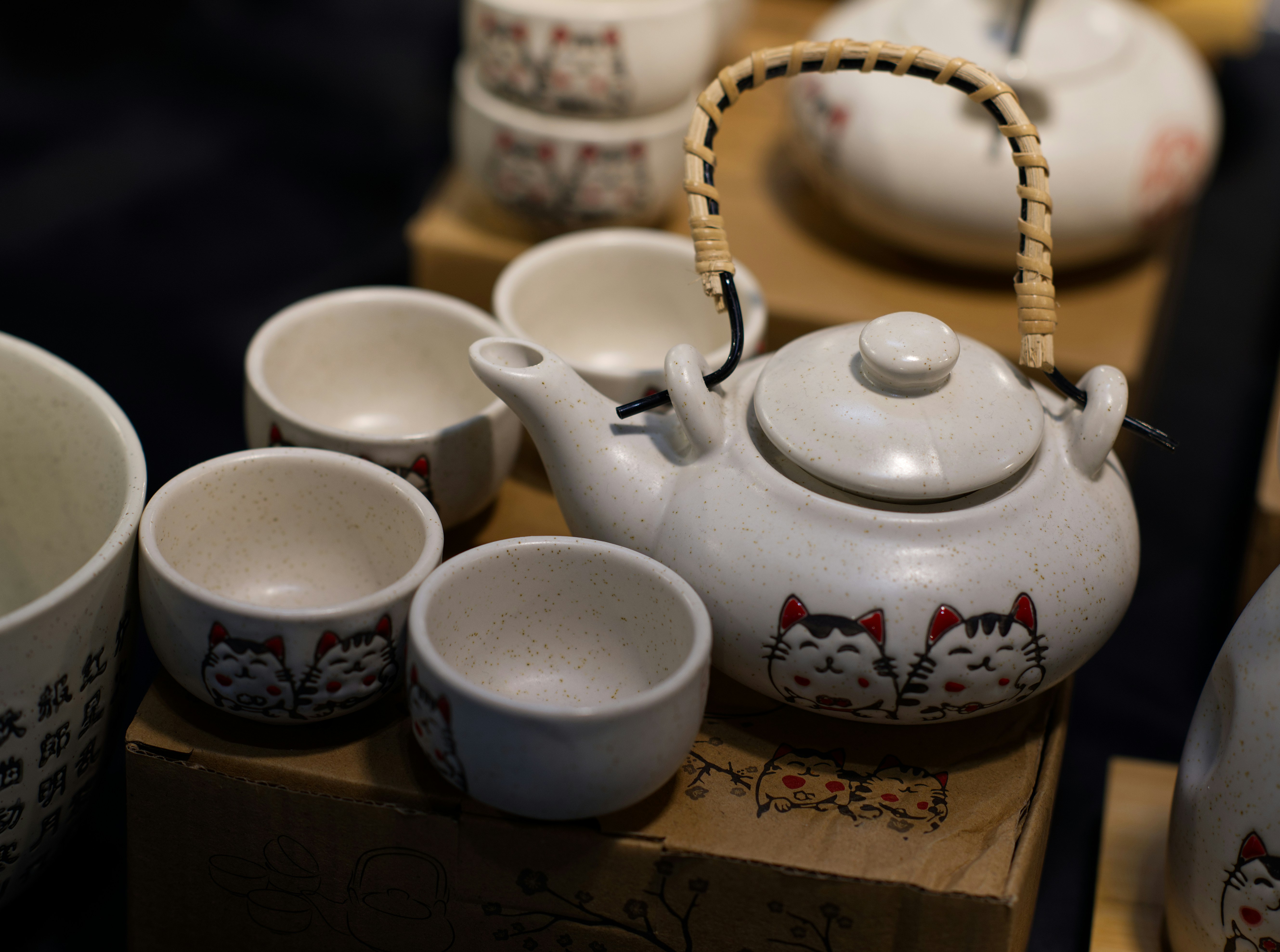 White ceramic teapot with a light-brown woven handle sits among small matching cups adorned with cat illustrations, arranged on a cardboard surface. A cozy still-life featuring delicate Japanese-style ceramics.