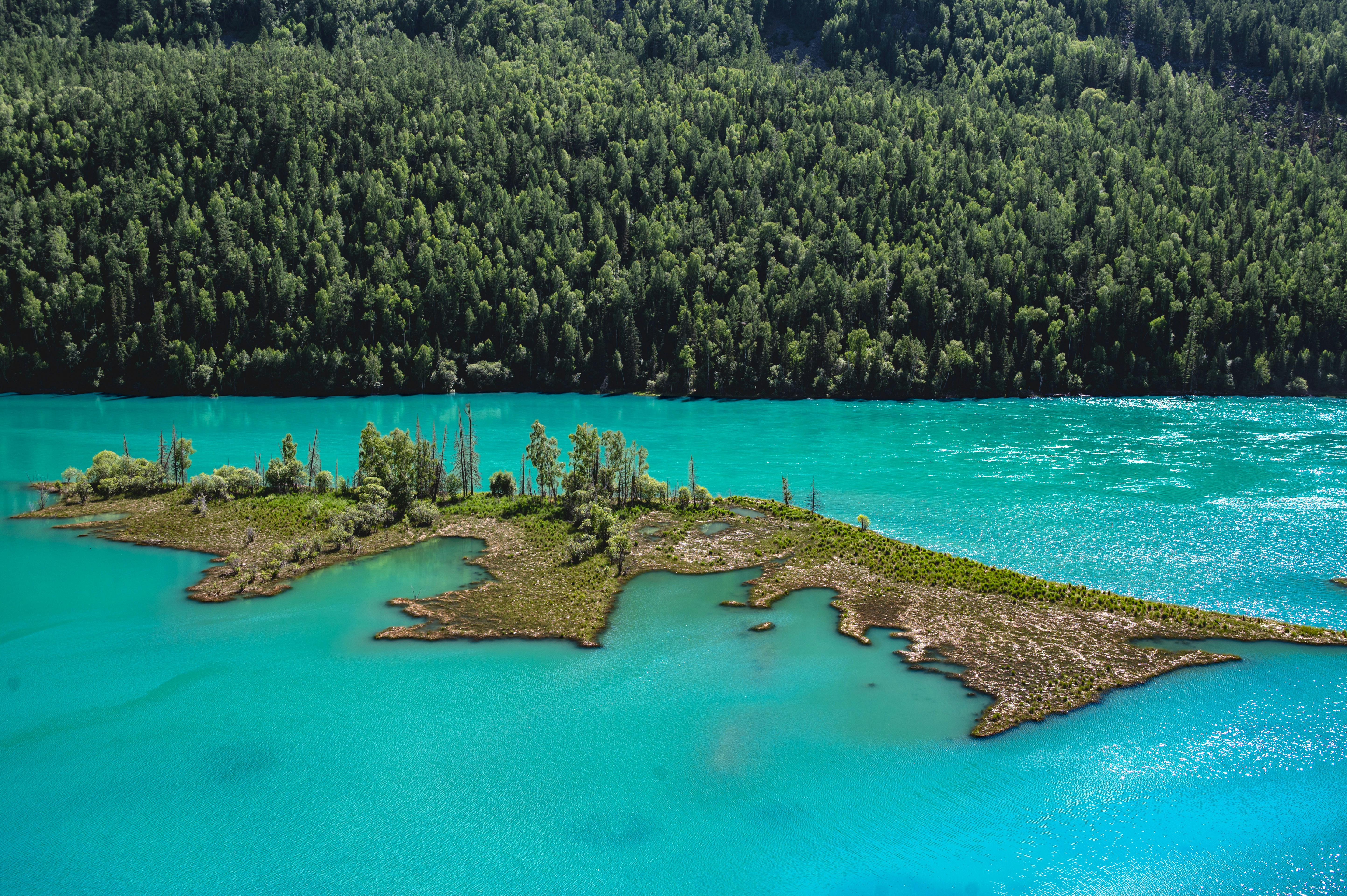Emerald islet surrounded by turquoise water, with a forested shoreline in the background and a narrow, sculpted landform extending into a shallow bay.