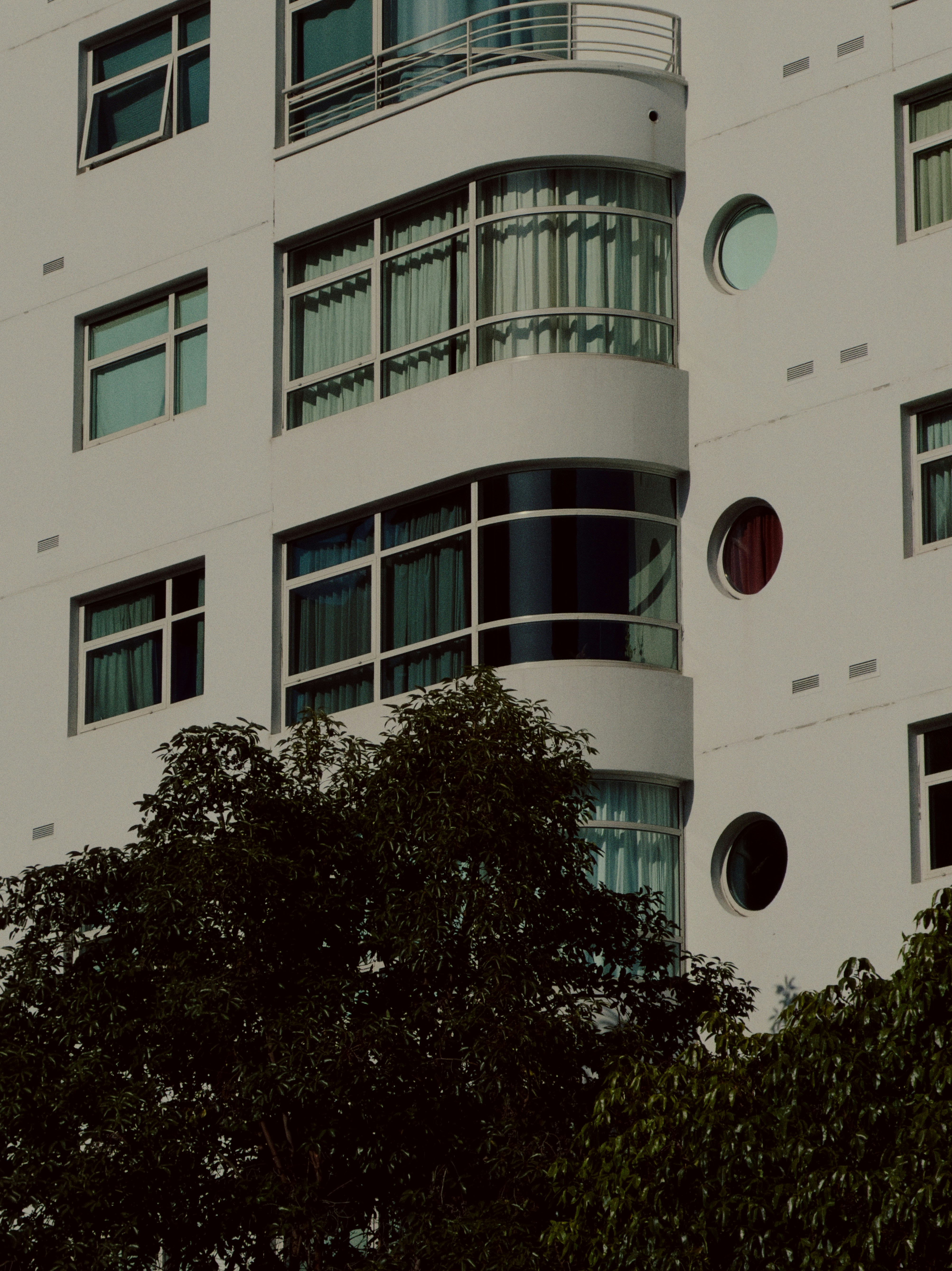 A white apartment building with curved balconies and circular windows behind dark green trees