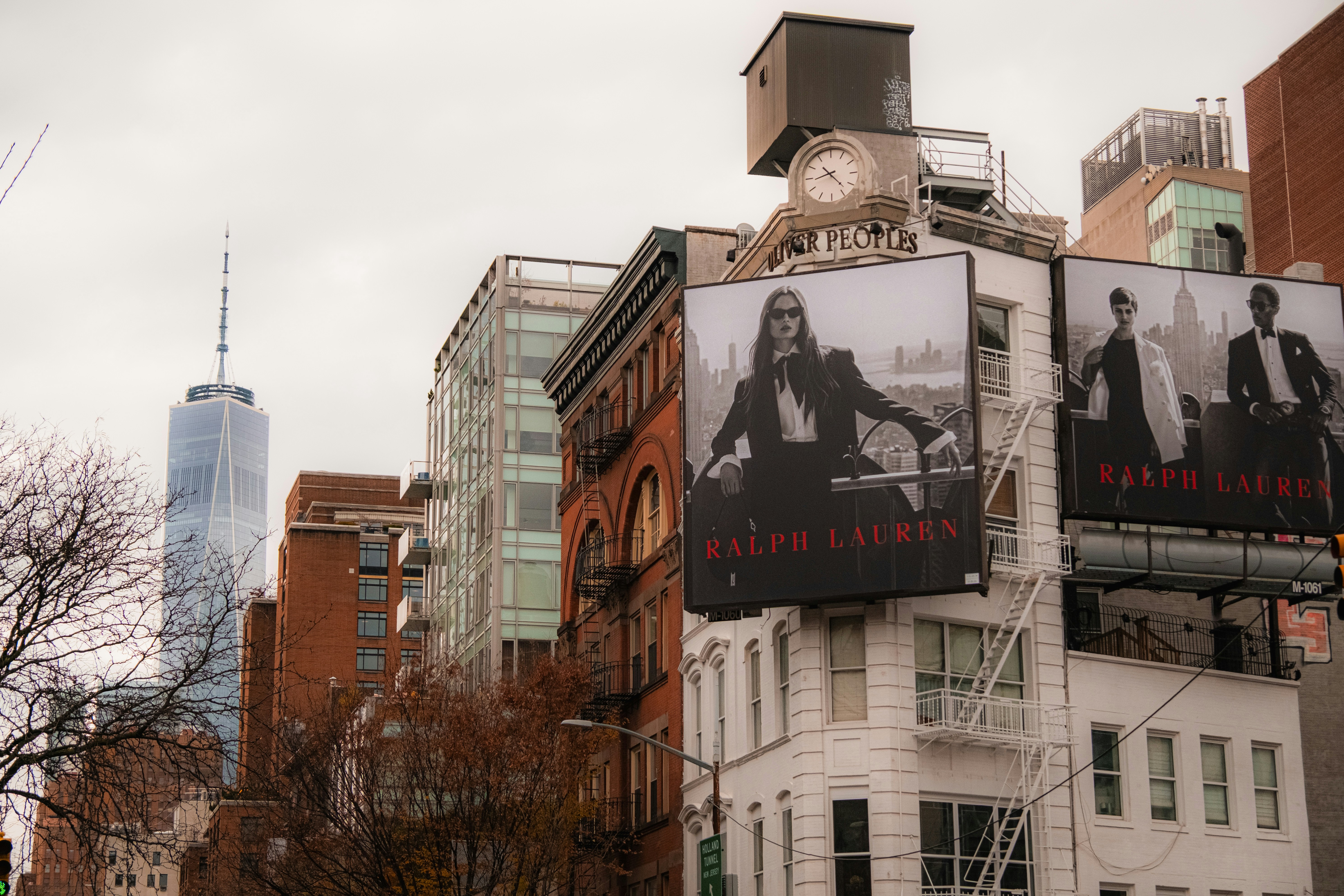 Billboard-heavy cityscape with Ralph Lauren ads on a historic building front, framed by leafless trees and distant skyscraper.