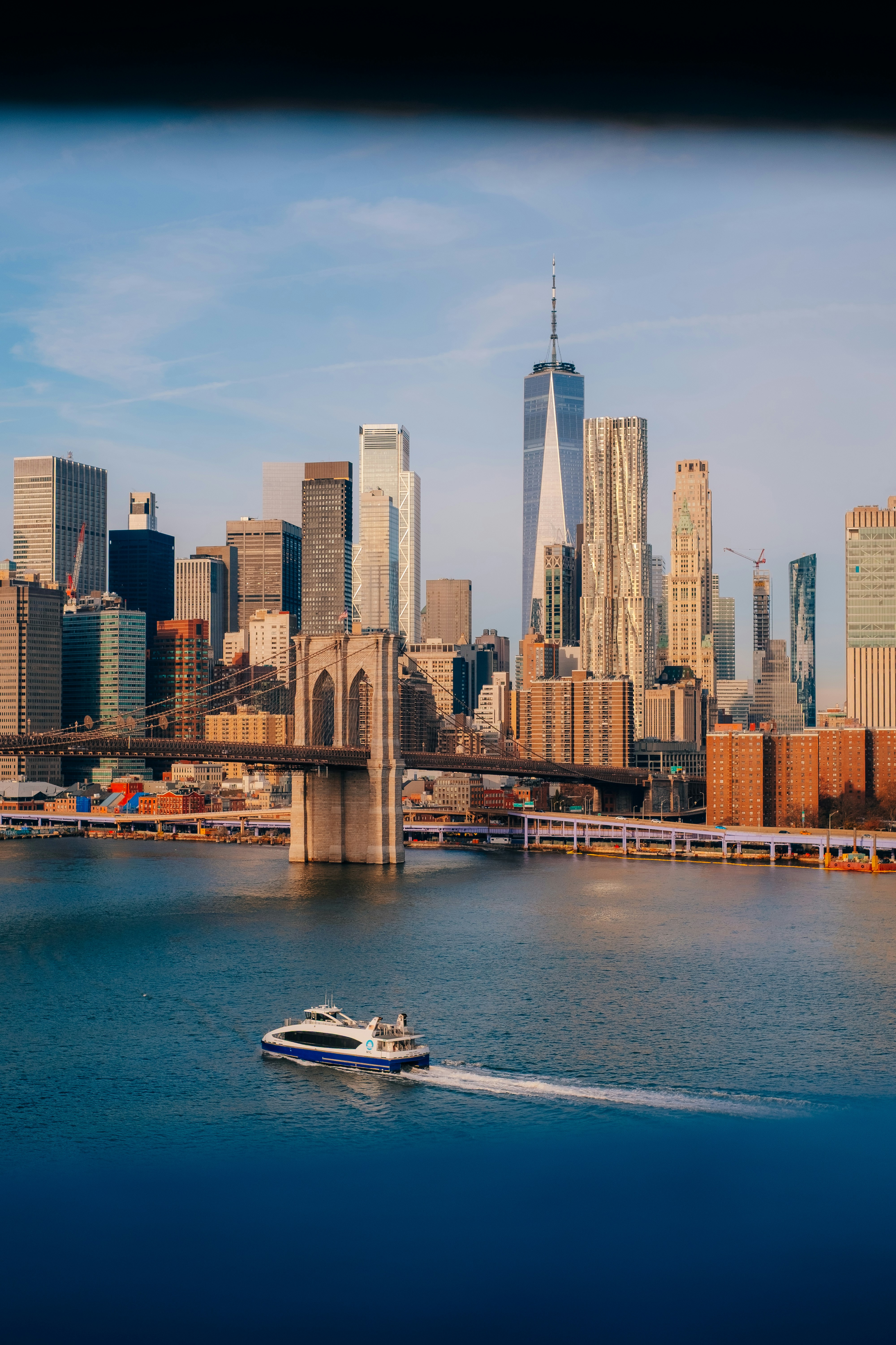 Sunlit skyline with the Brooklyn Bridge and a ferry gliding across the East River under a clear sky. A tall, iconic cluster of skyscrapers rises in the background, showcasing urban energy and river life.