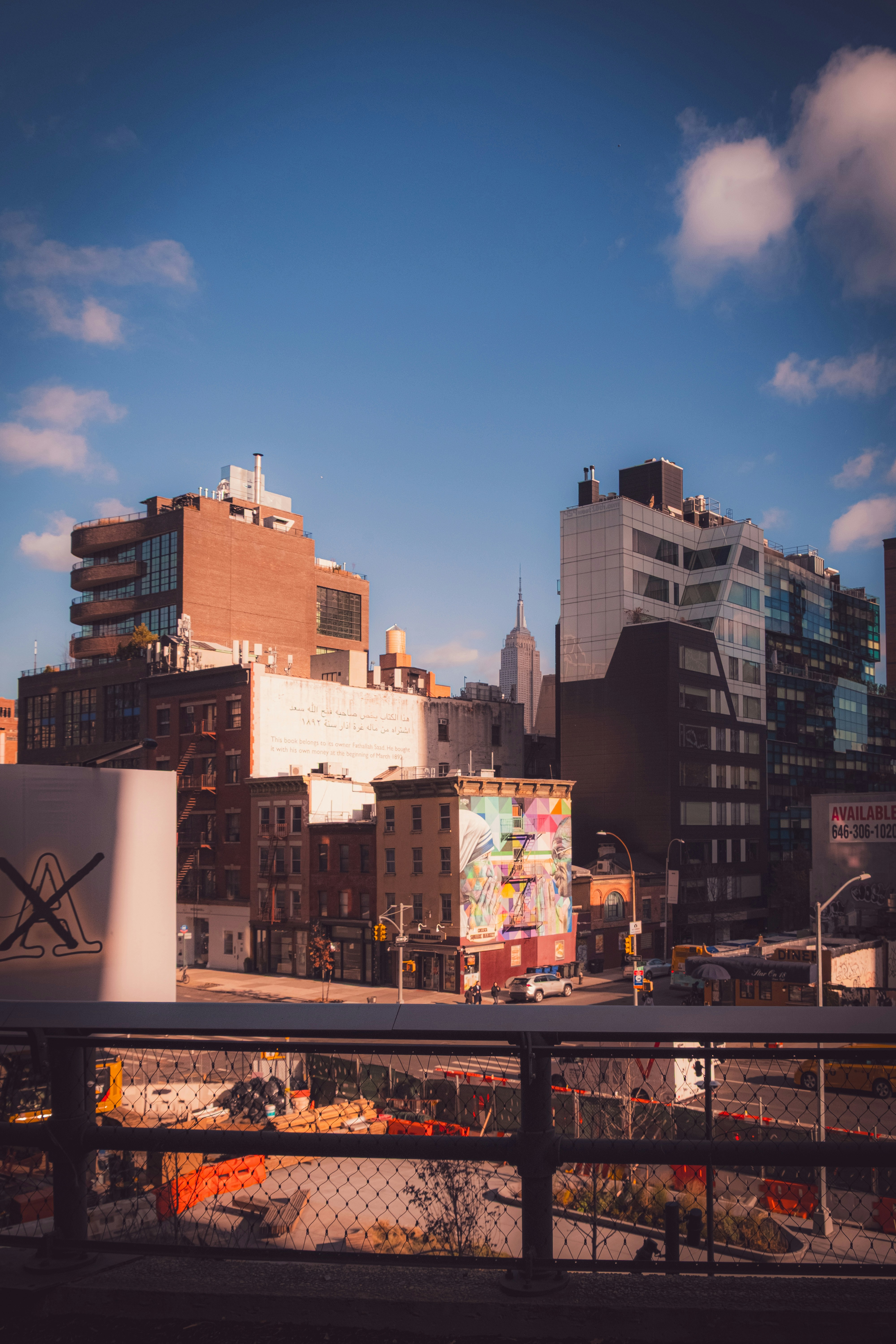 Skyline with modern buildings bathed in late-day light; foreground shows a railing and construction site beneath, suggesting urban redevelopment.