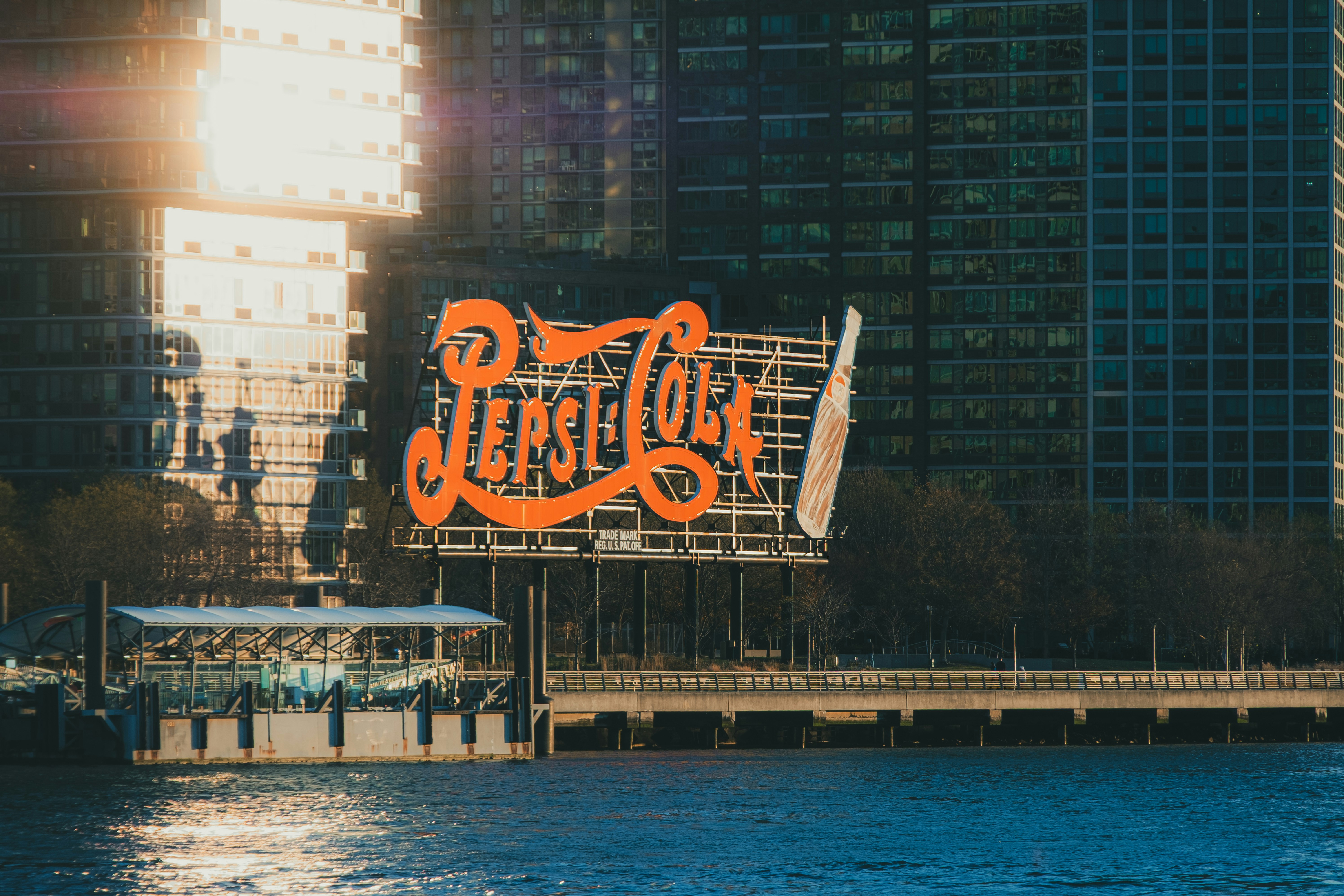 Bright orange sign atop a riverfront structure against dark glass buildings, reflecting on the water; a hint of urban nightlife and modern architecture.