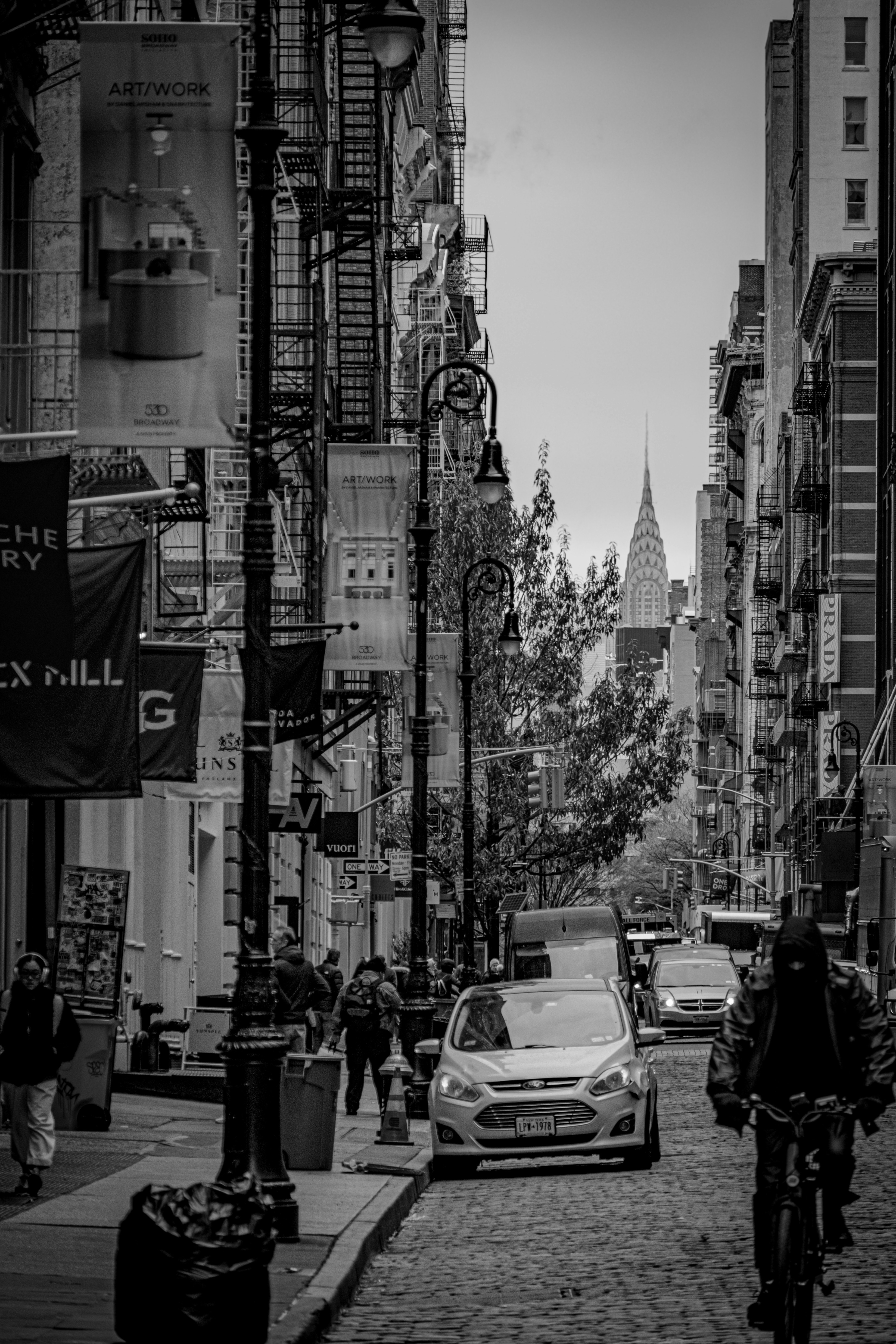 A grayscale city street scene with pedestrians, a car, and a cyclist along a cobbled road; architectural details line both sides as a distant spire rises above the skyline.