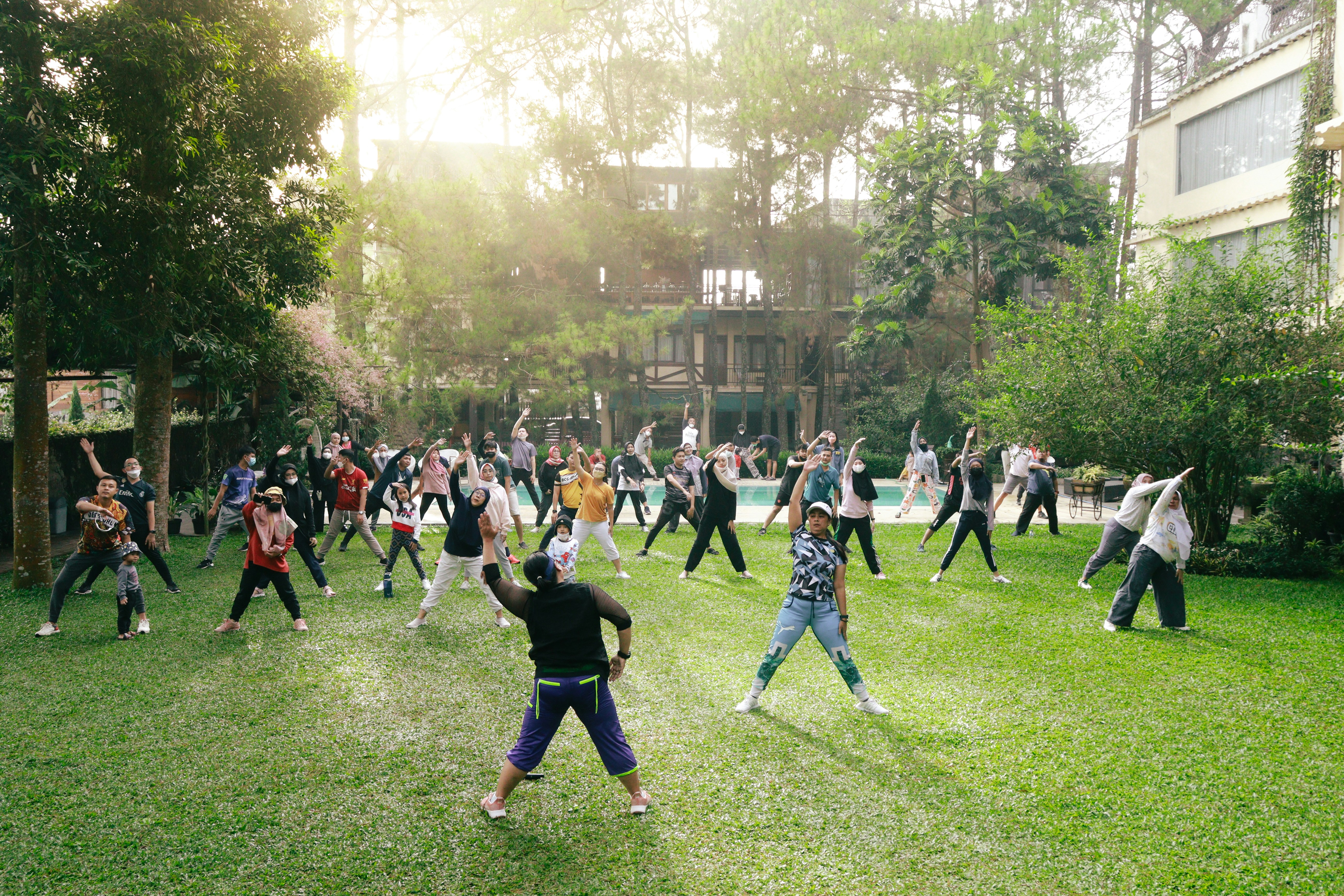 A large outdoor group dance class on a sun-dappled campus lawn, with participants arranged in staggered rows and arms raised in motion.