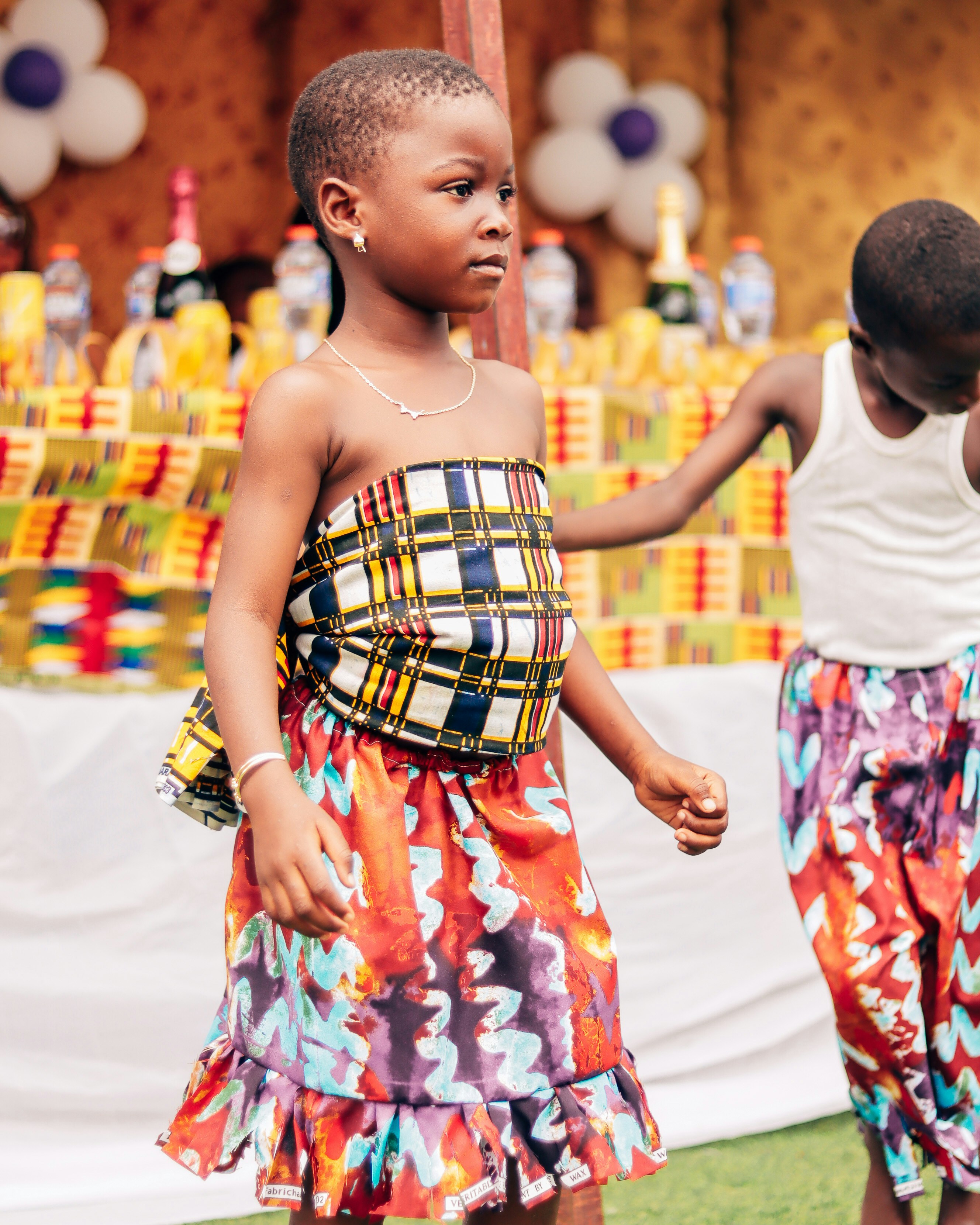 Sunlit Parade: A Young Girl in Colorful Traditional Attire Strolls at a Festive Market