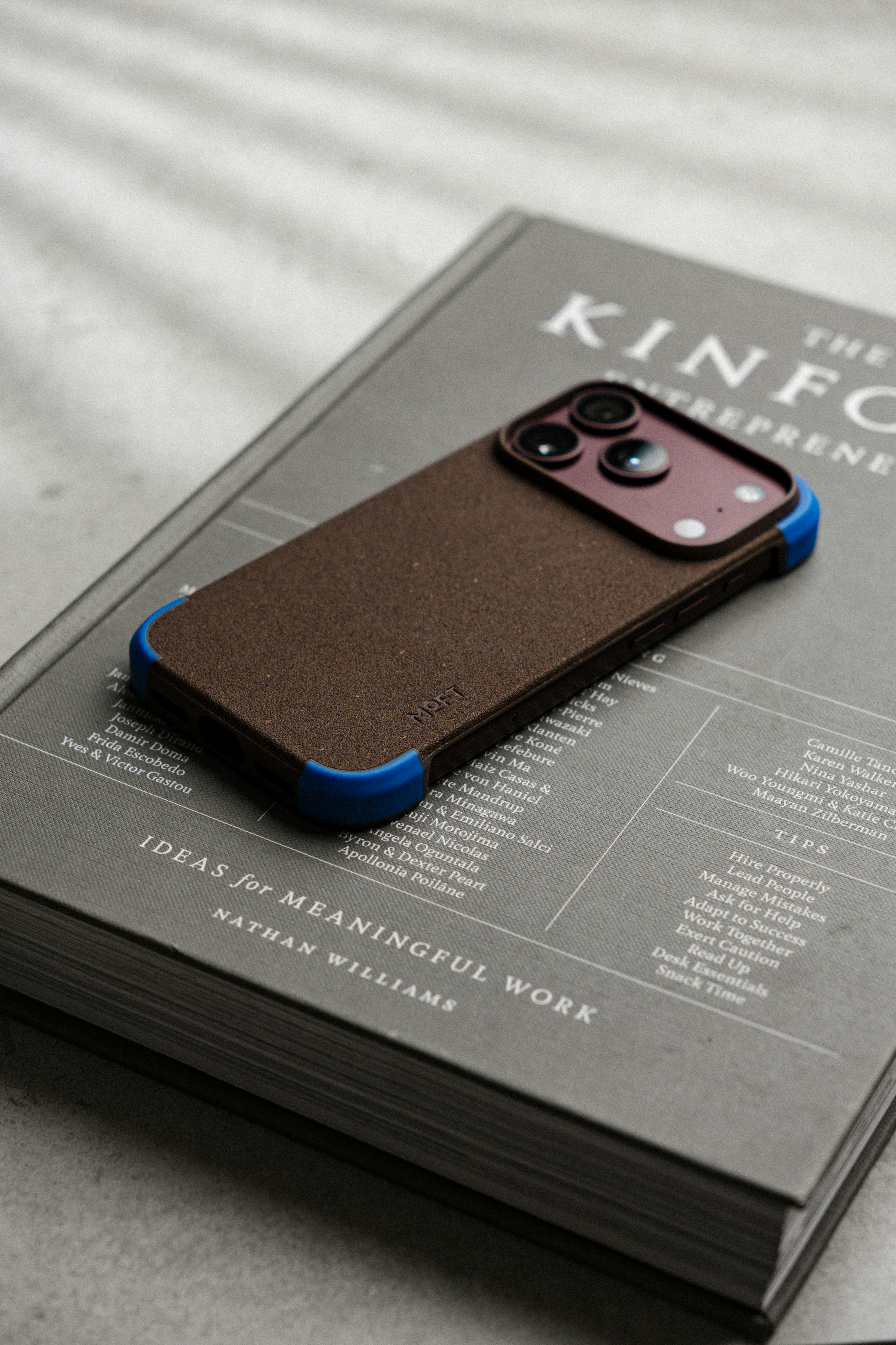 A brown phone with blue bumper sits on a dark book titled Kinfolk, highlighting a simple, modern workspace setup.